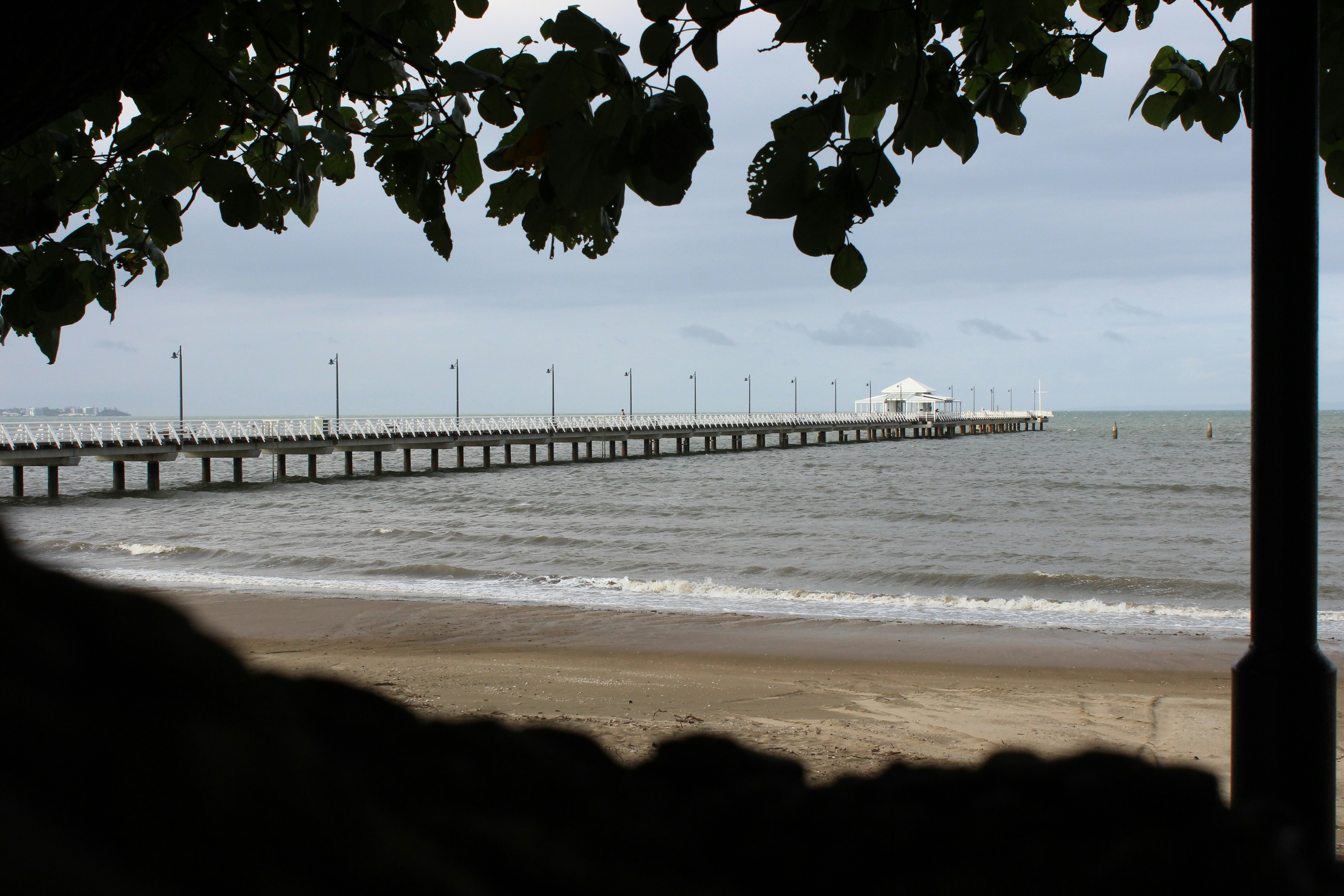 Long pier extending over calm sea, framed by leafy branches, under a cloudy sky.