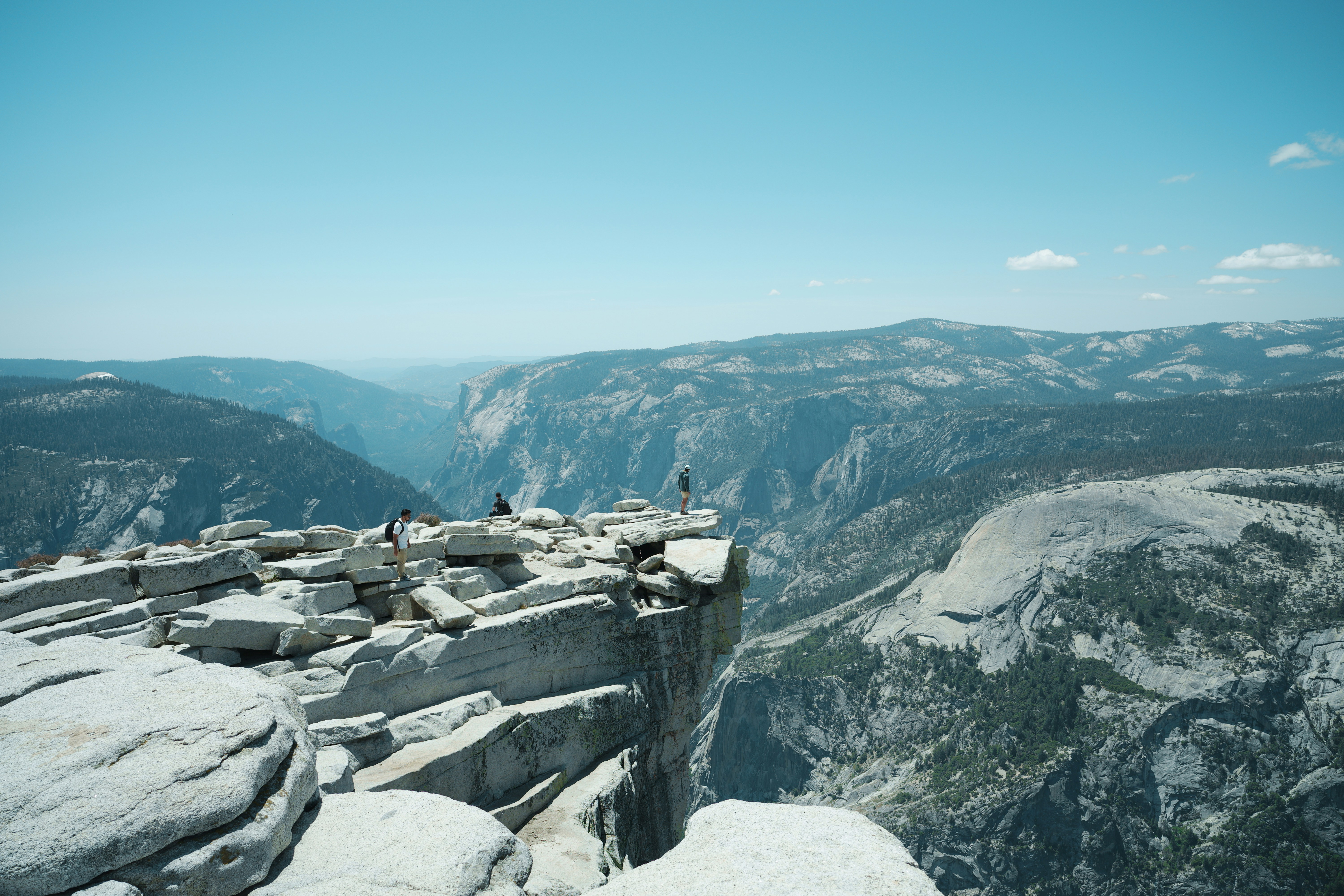 Hikers standing atop rocky cliffs with expansive mountain ranges in the background under a clear blue sky.