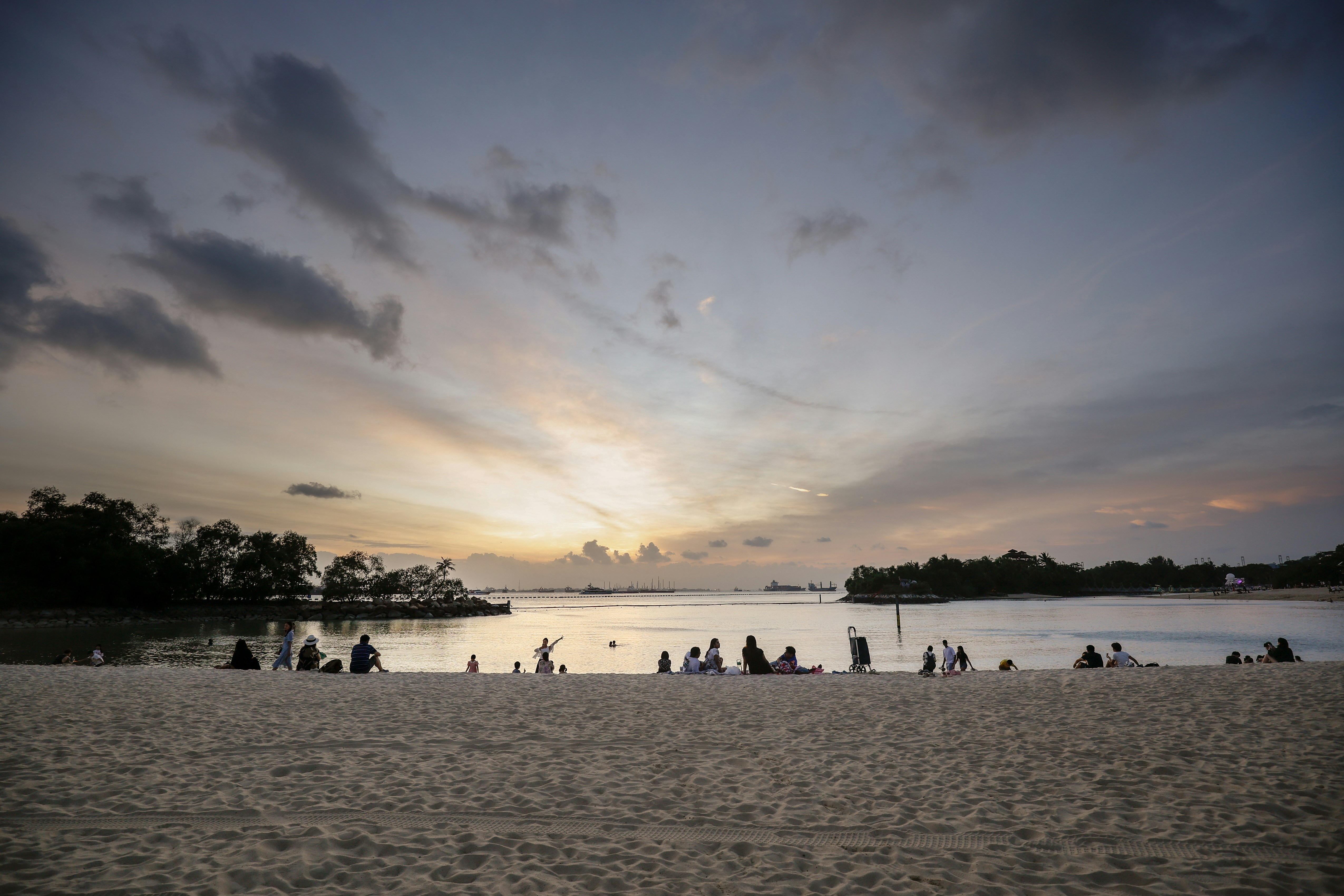 People gather on a sandy beach at dusk, silhouetted against a pastel sunset over calm waters.