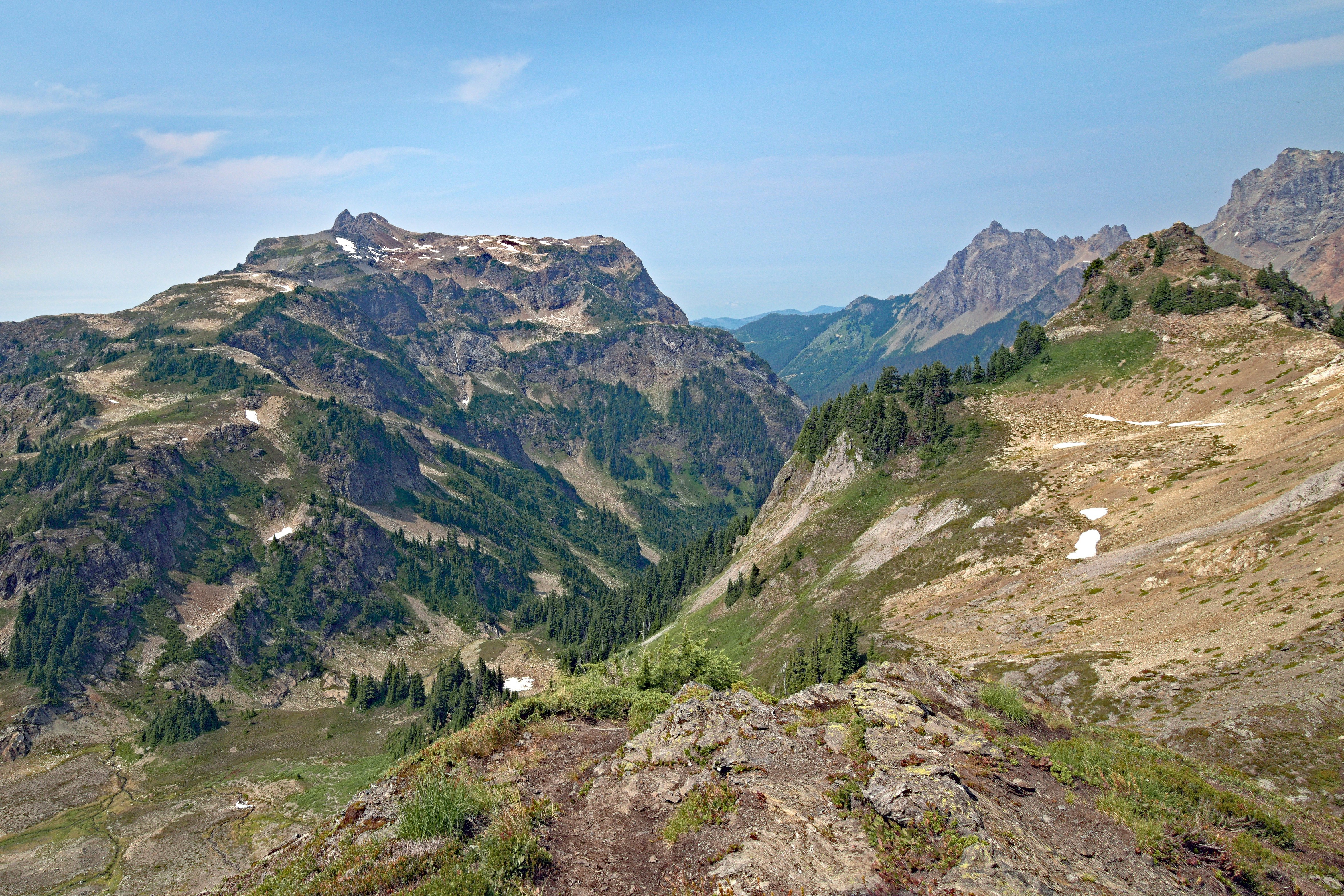 Mountainous landscape with rugged peaks, green valleys, and patches of snow under a clear blue sky.