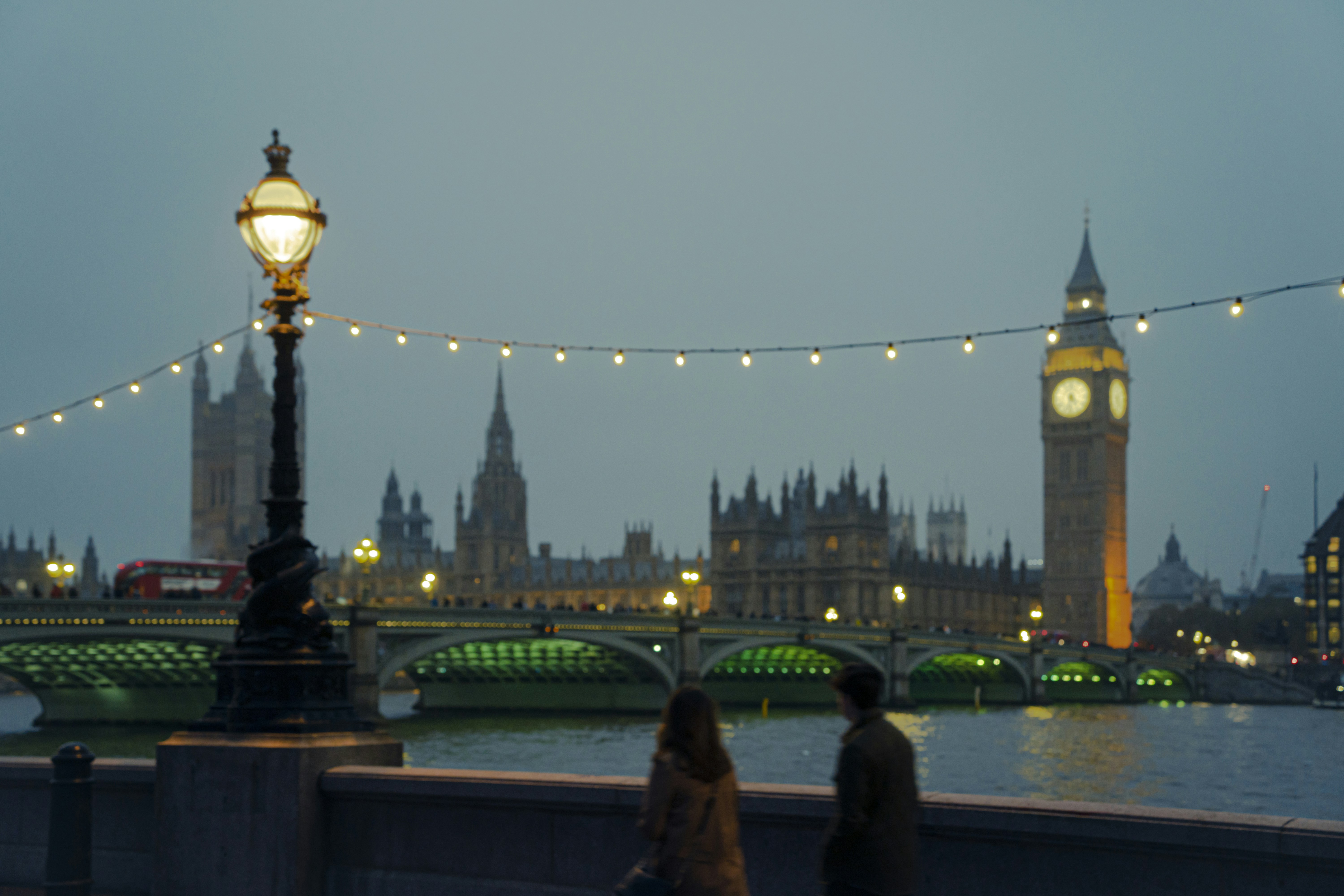 Evening view of Westminster Bridge and Big Ben under string lights, with silhouettes of two people walking.