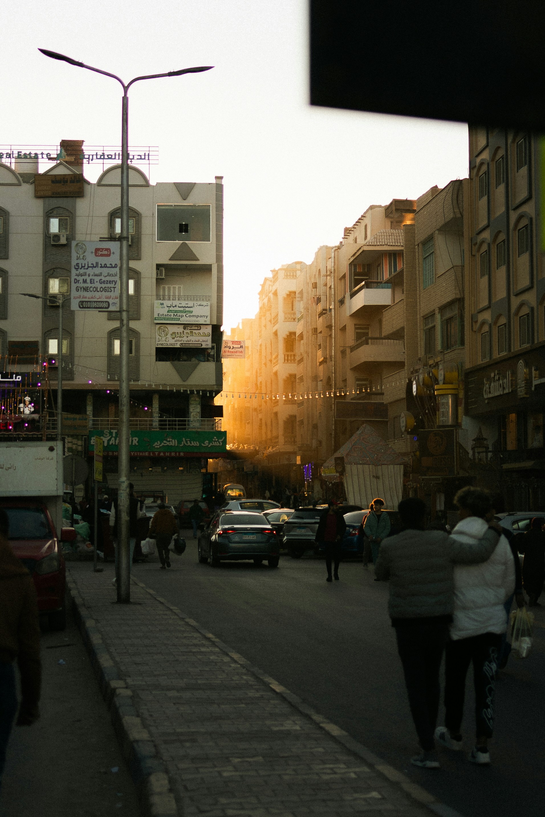 Narrow city street bathed in warm sunset light, with people walking and cars lining the road.