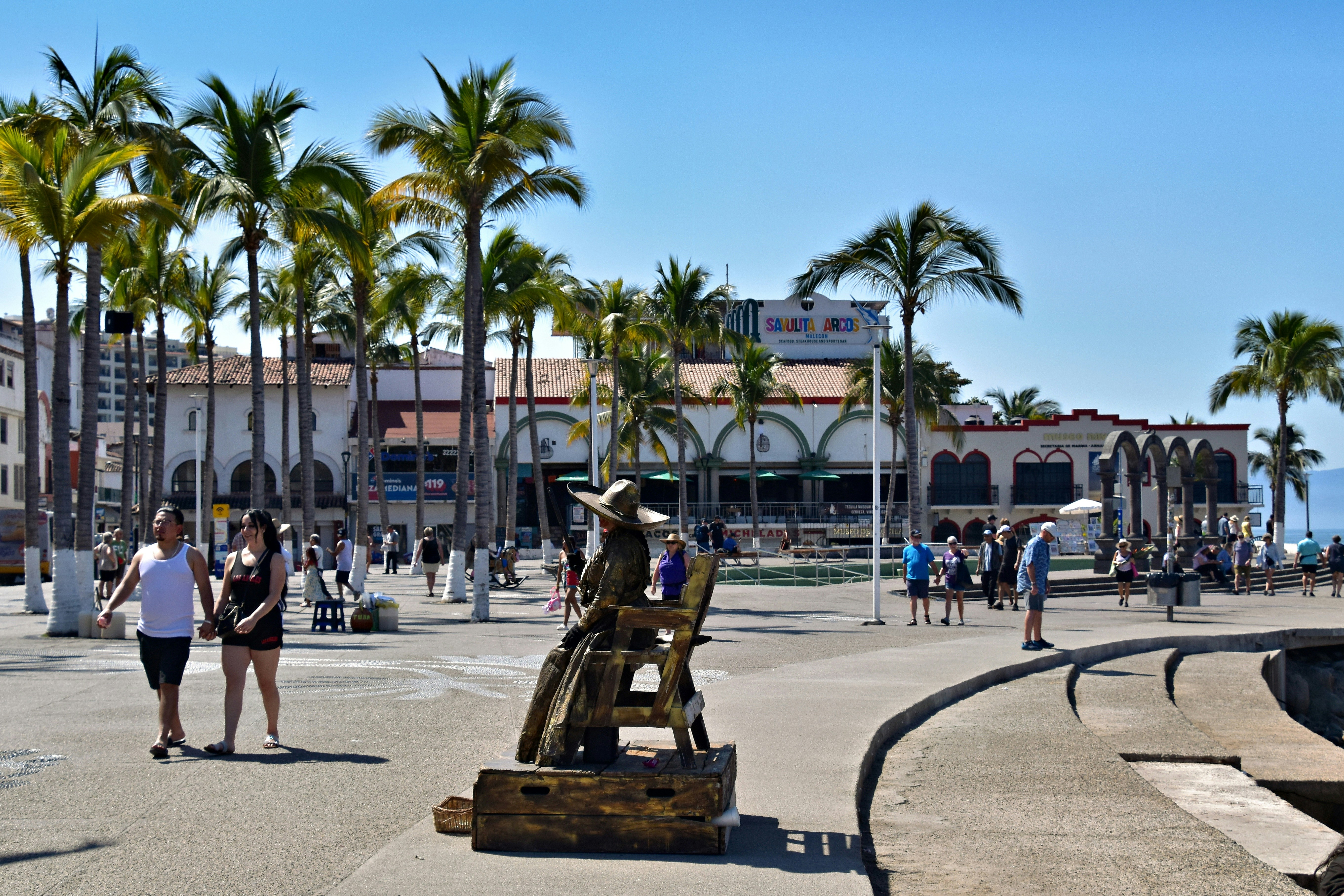 People walking along a palm-lined promenade with a bronze statue in the foreground under a clear blue sky.