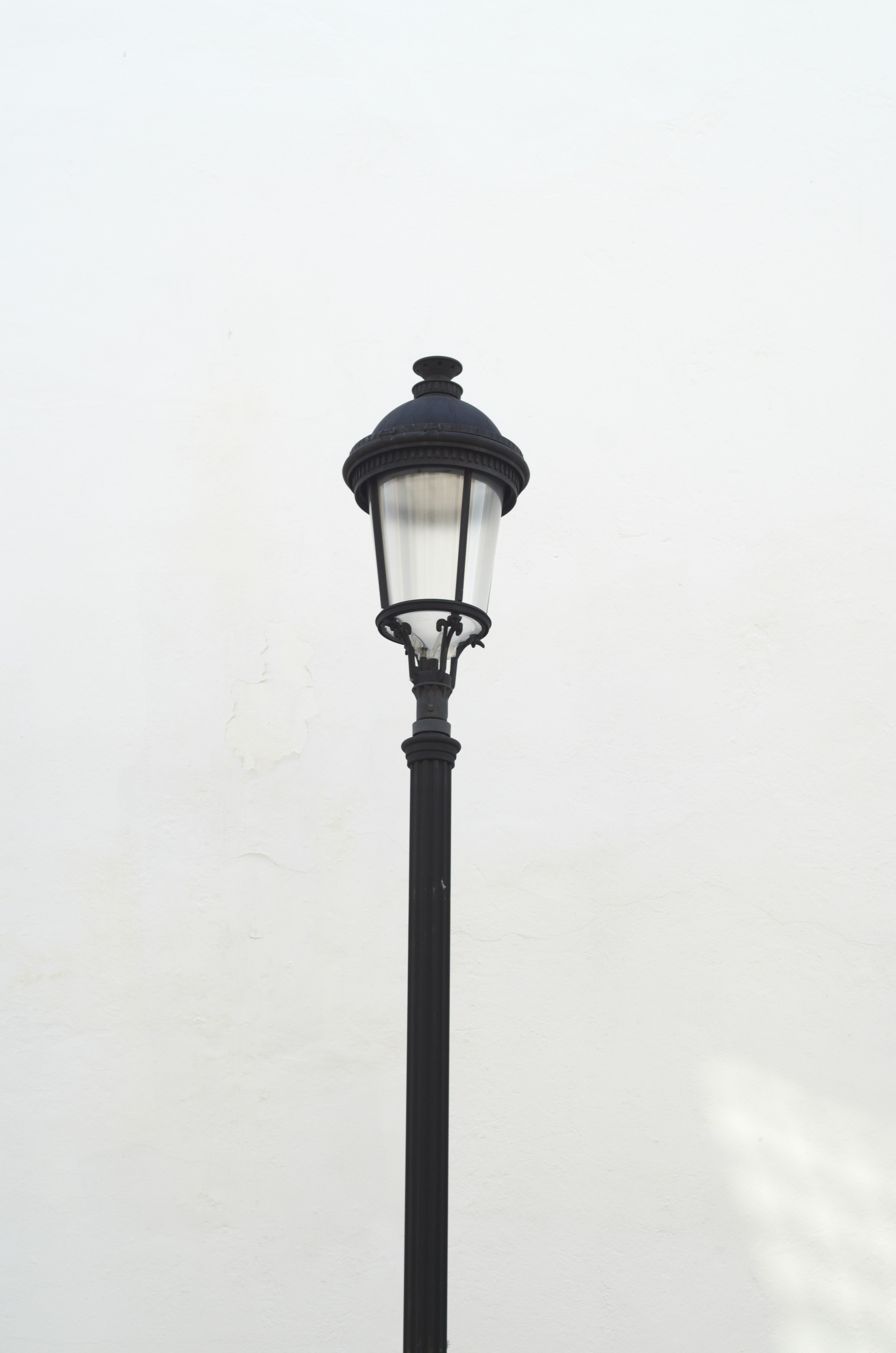 A black street lamp rises from a tall post against a pale wall. The glass lantern catches soft light, emphasizing its classic silhouette.