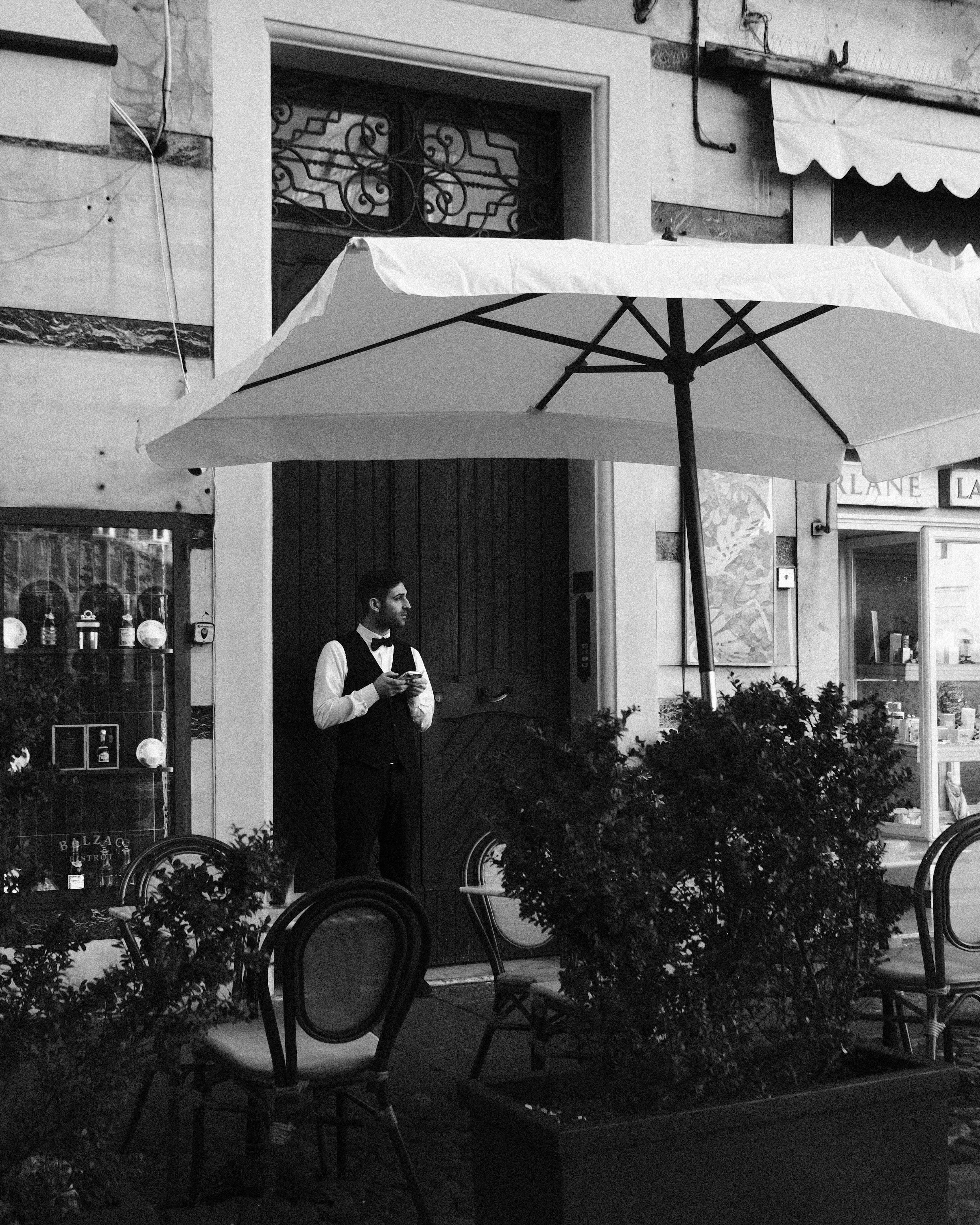 Beneath the Café Umbrella: a Dapper Waiter Stands Quietly at the Threshold
