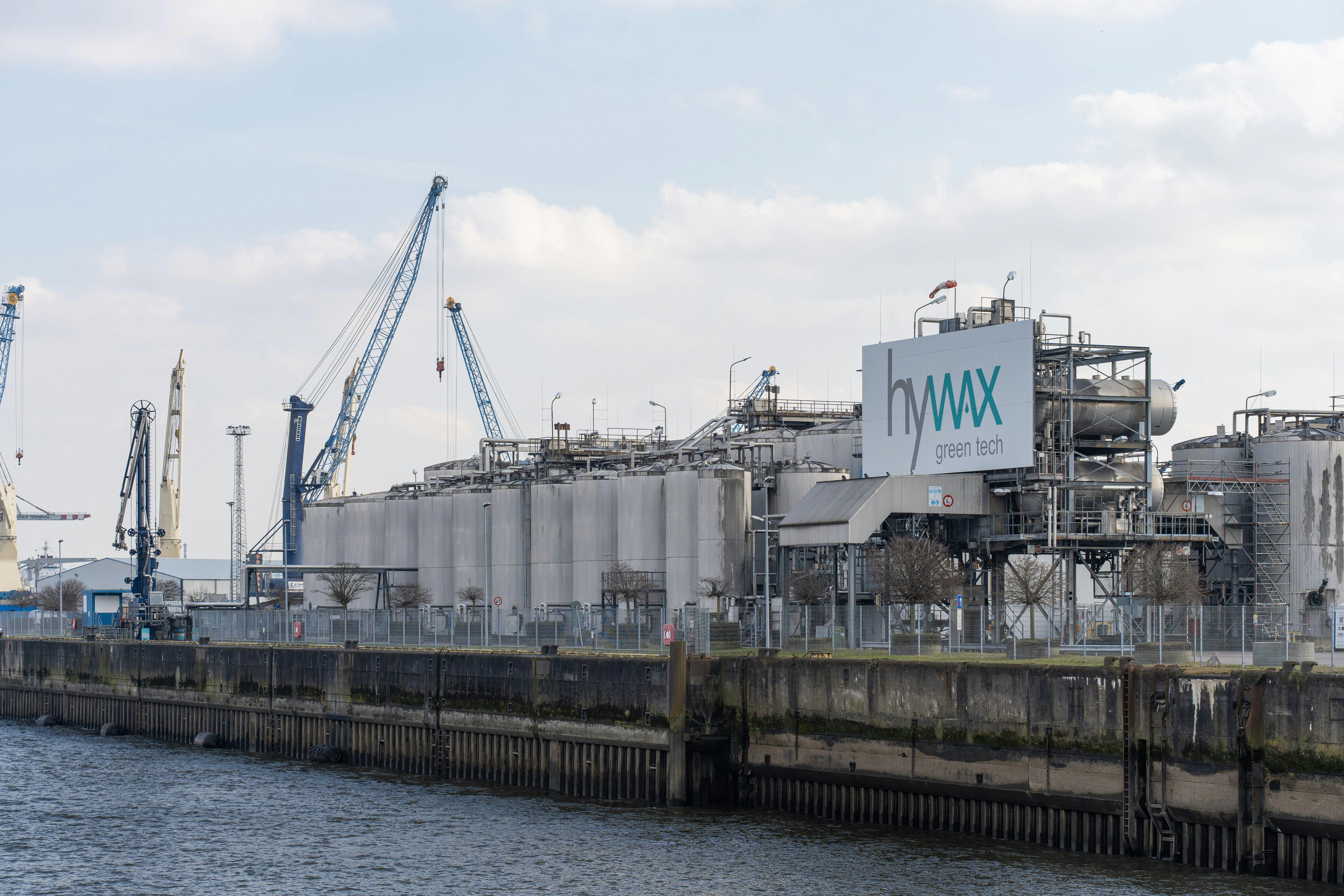 Industrial complex with cranes and buildings along a waterfront under a partly cloudy sky.