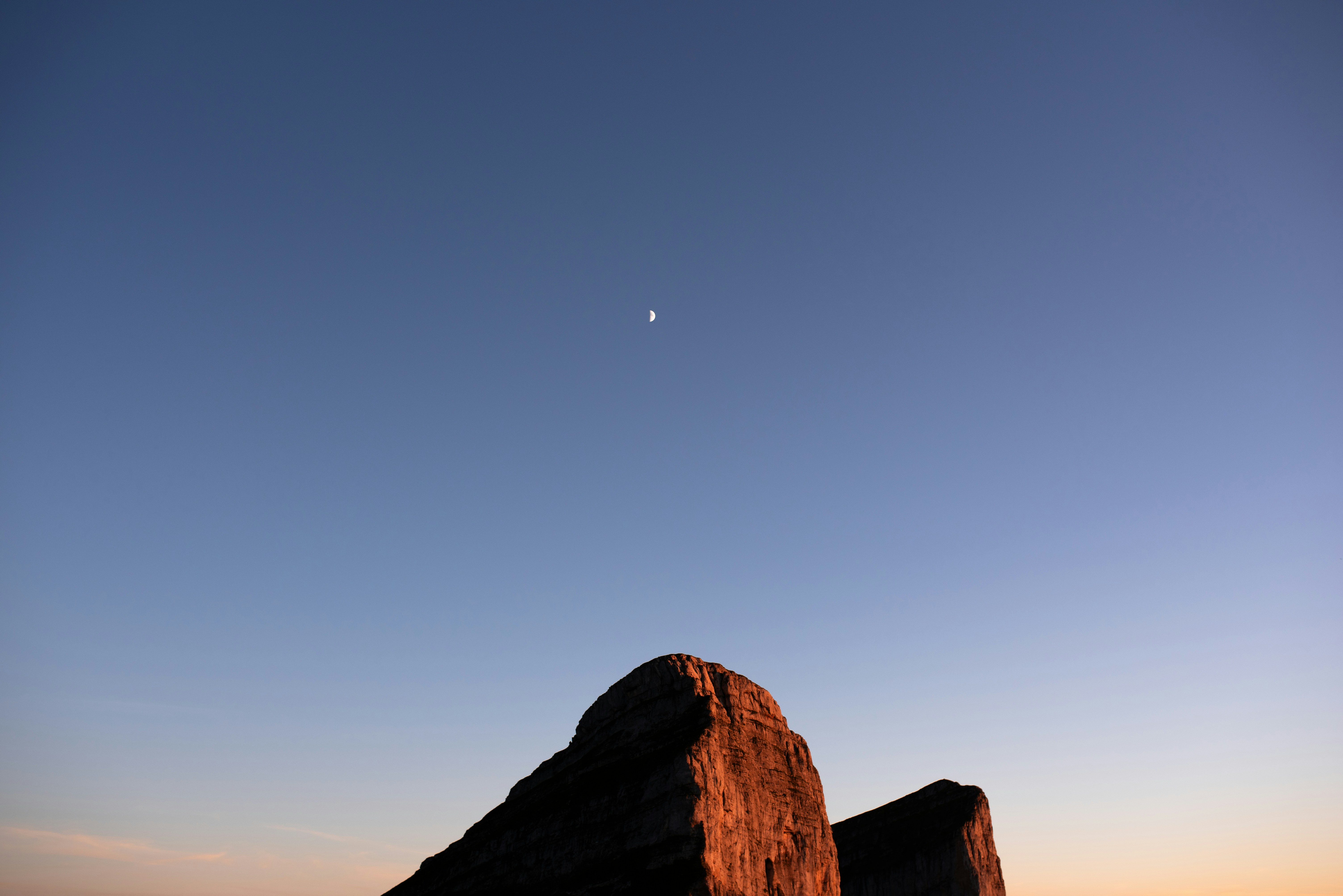 Dusky sky with a crescent moon above sunlit mountain peaks at sunset.