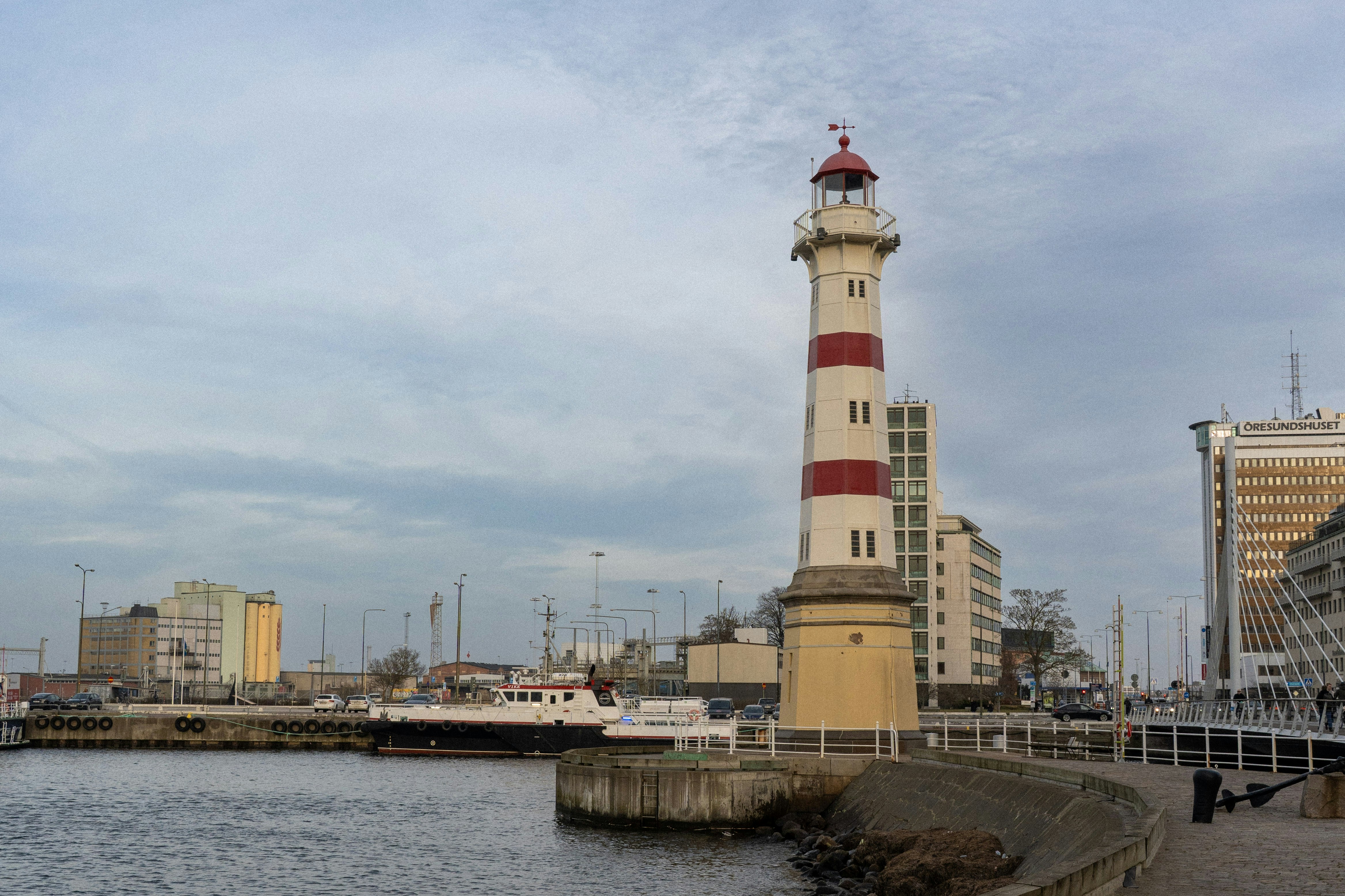 Striped lighthouse stands by a city waterfront with buildings and boats in view.