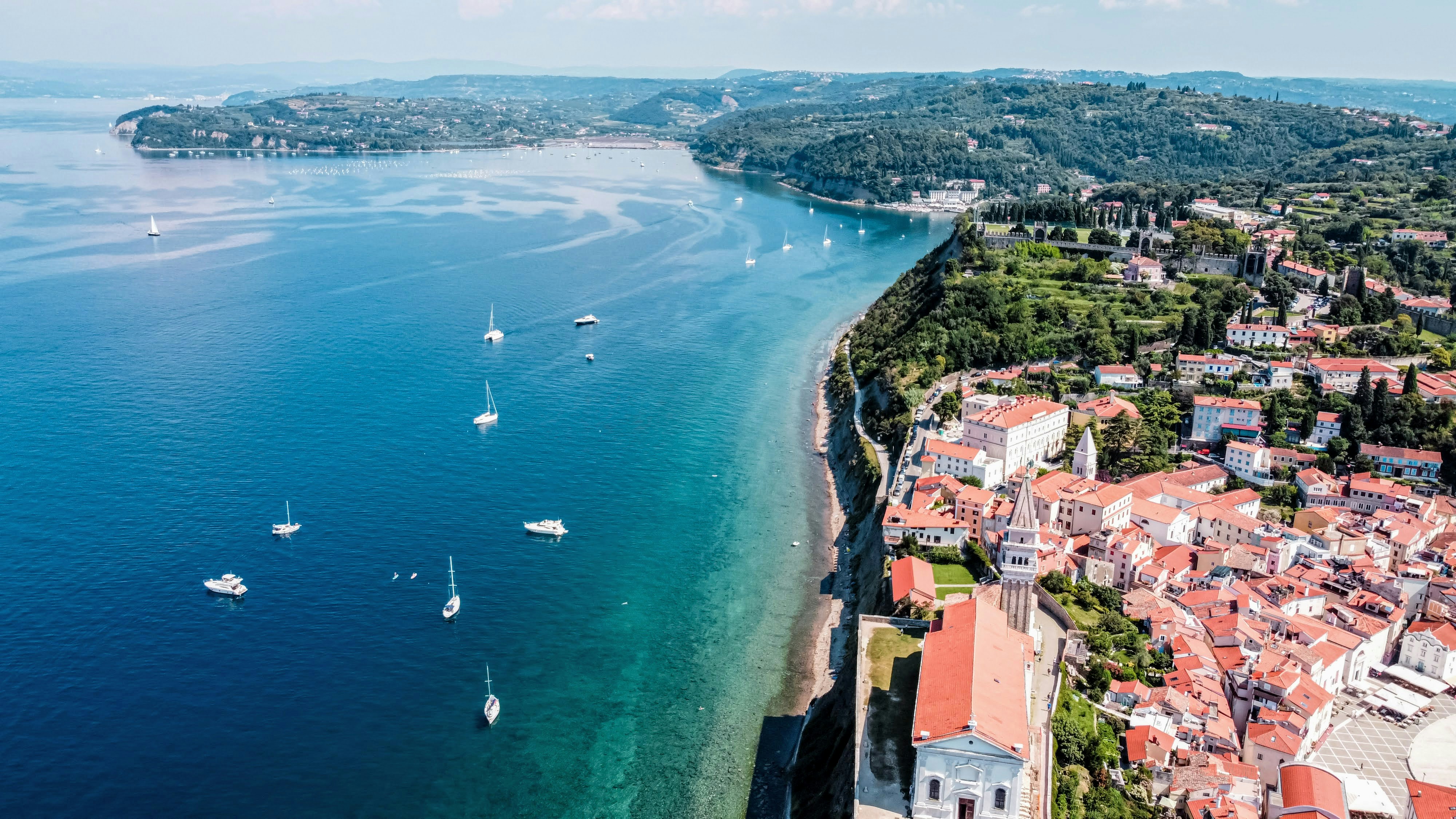 Aerial view of a coastal town with terracotta rooftops beside a deep blue sea dotted with white sailboats.