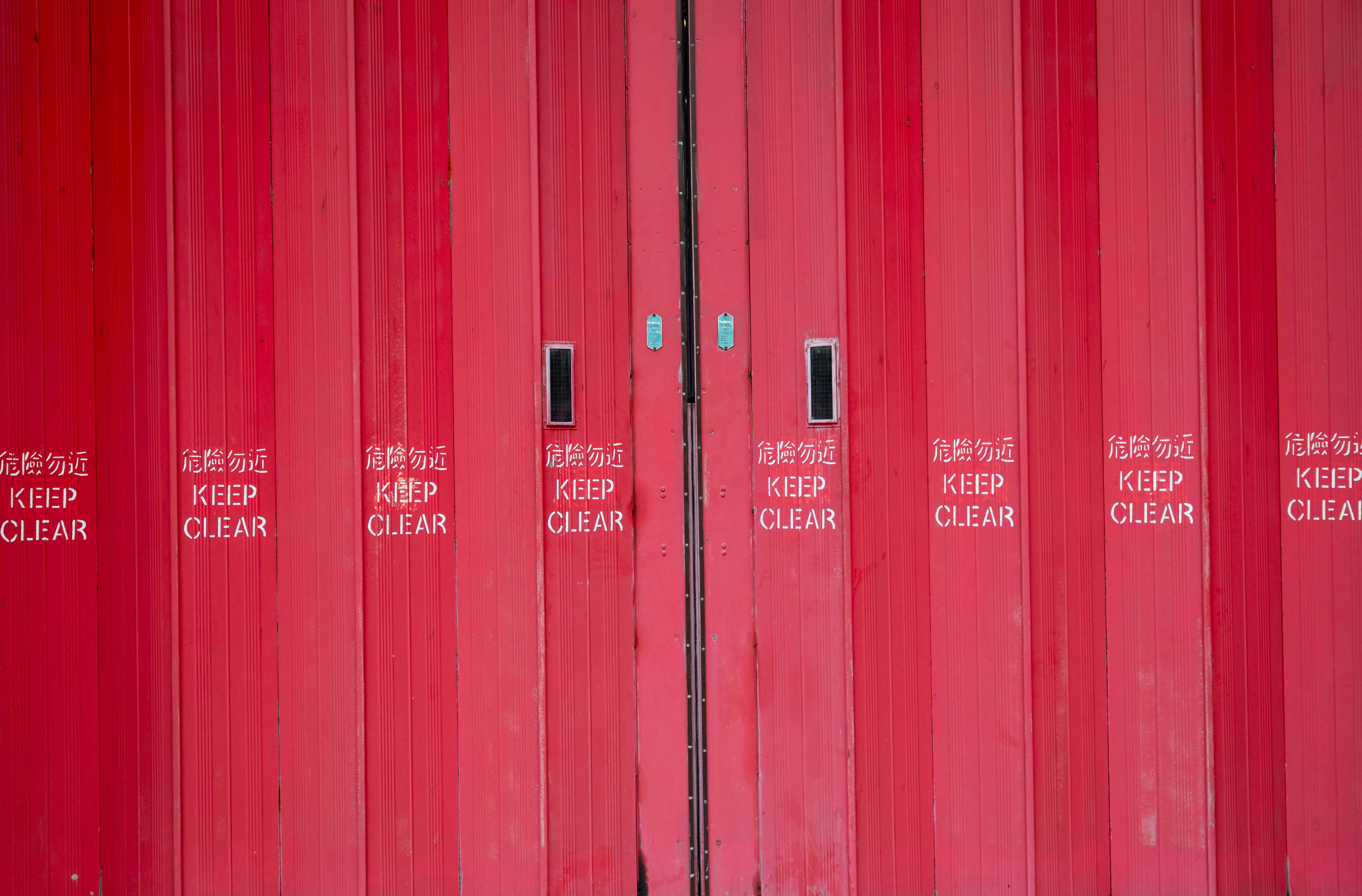 A bold red set of fire station doors dominates the frame, featuring repeated "KEEP CLEAR" warnings in both English and Chinese. The industrial design, with its uniform lines and symmetry, emphasizes order and function. The vivid color and stark lettering serve as a clear directive, symbolizing urgency and preparedness in an urban environment.