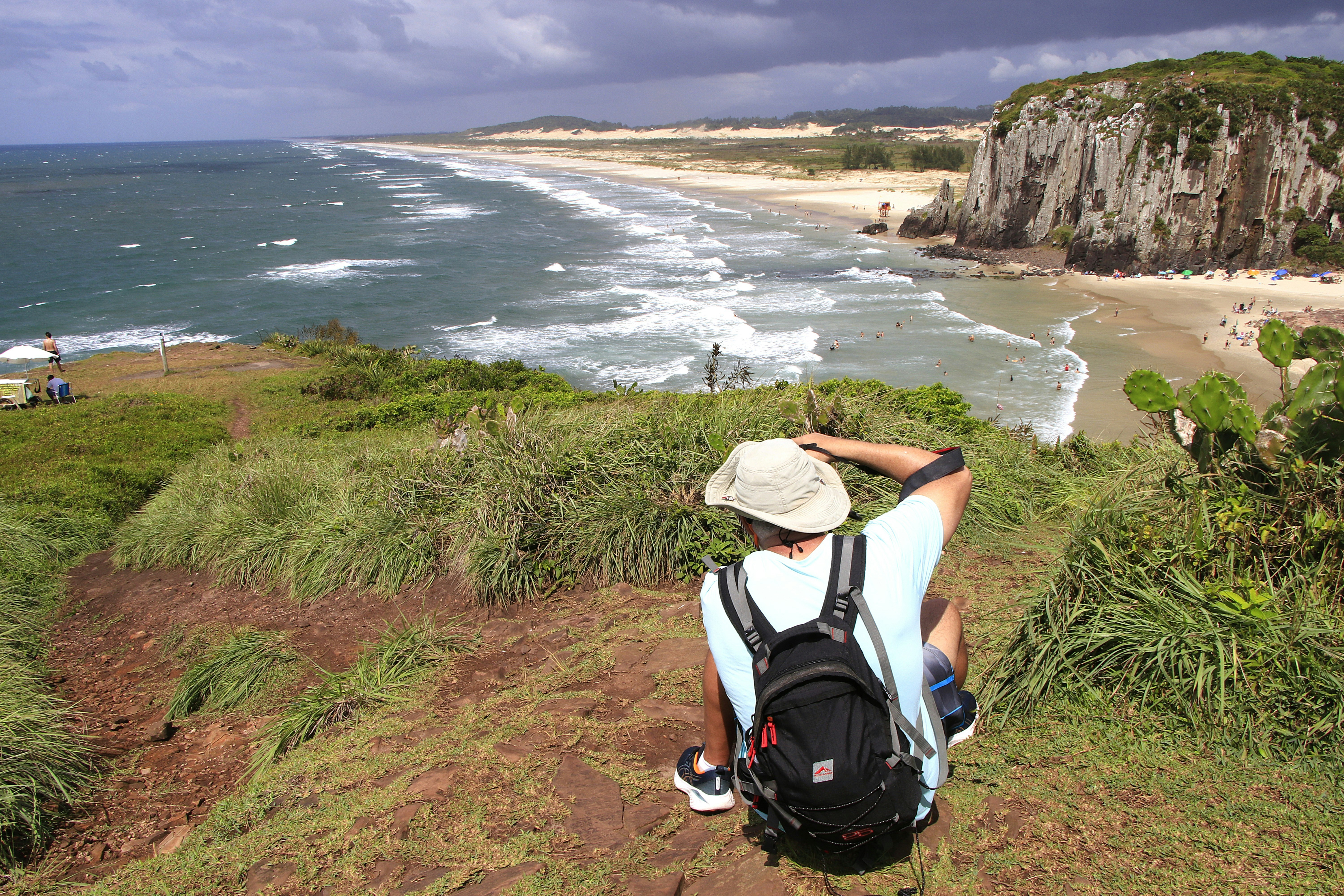 Person sitting on a grassy cliff overlooking a vast beach and ocean under a cloudy sky.