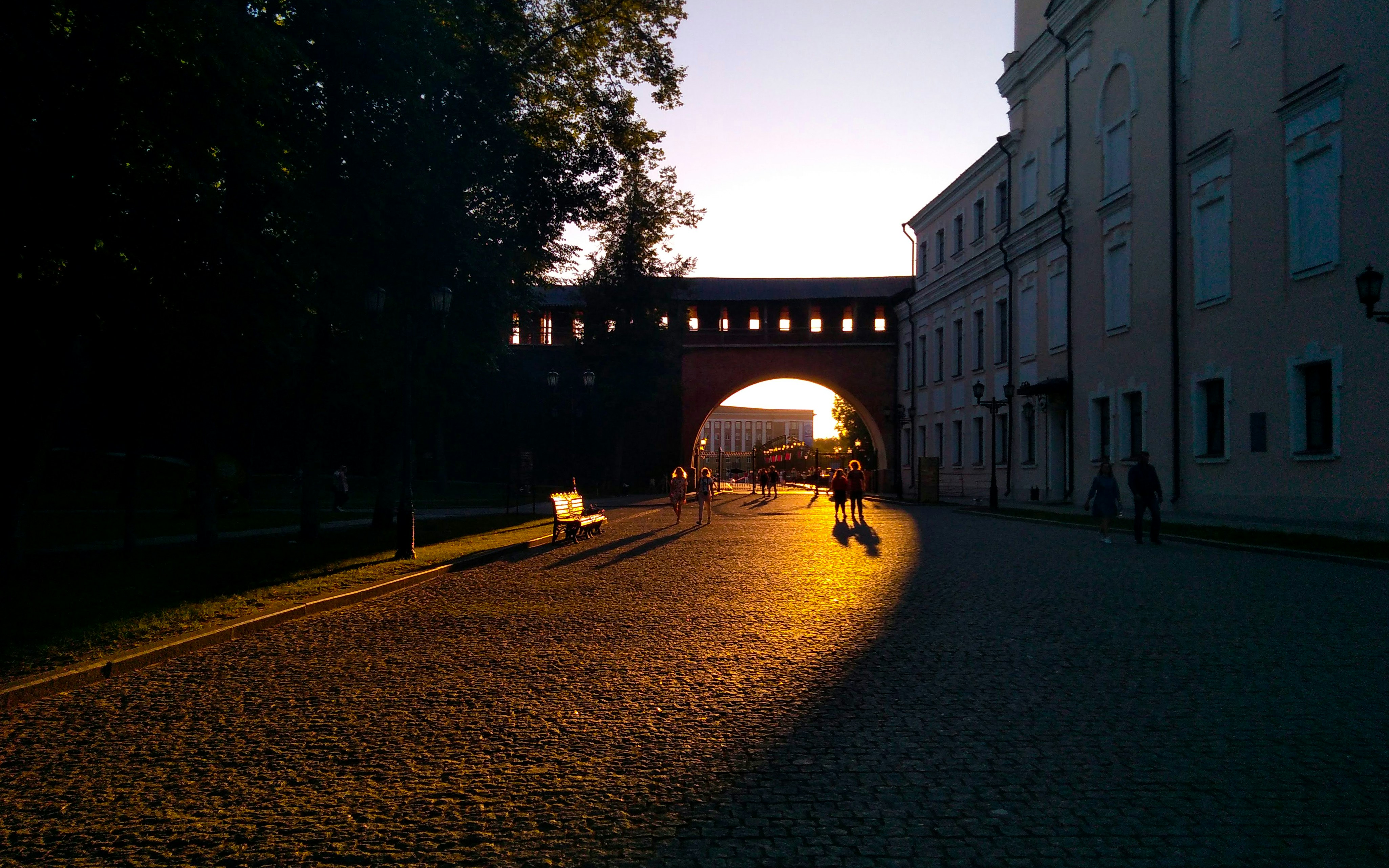 People walking under an archway with golden sunlight casting long shadows on cobblestone path.