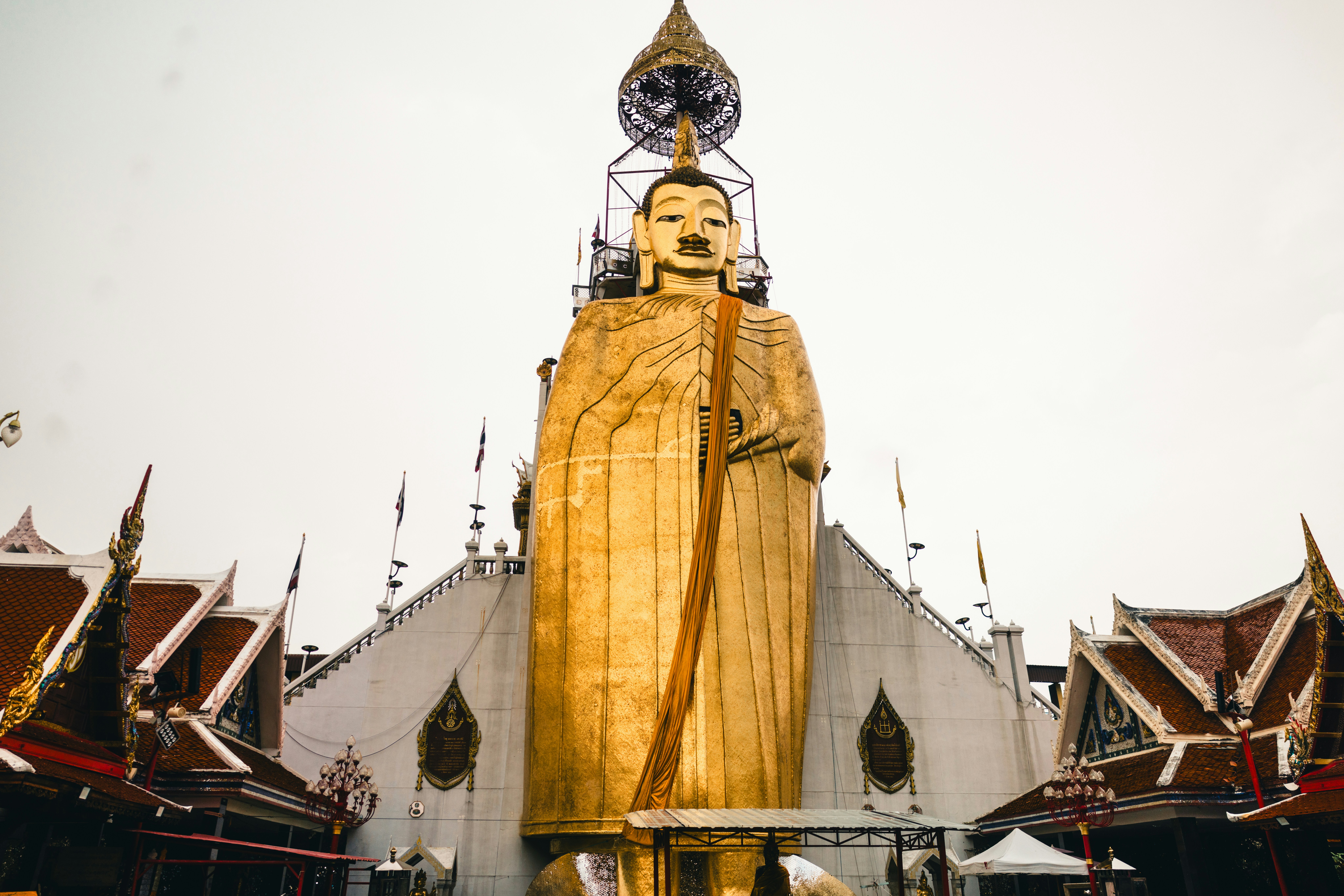Tall golden Buddha statue standing serenely against a backdrop of traditional Thai architecture.