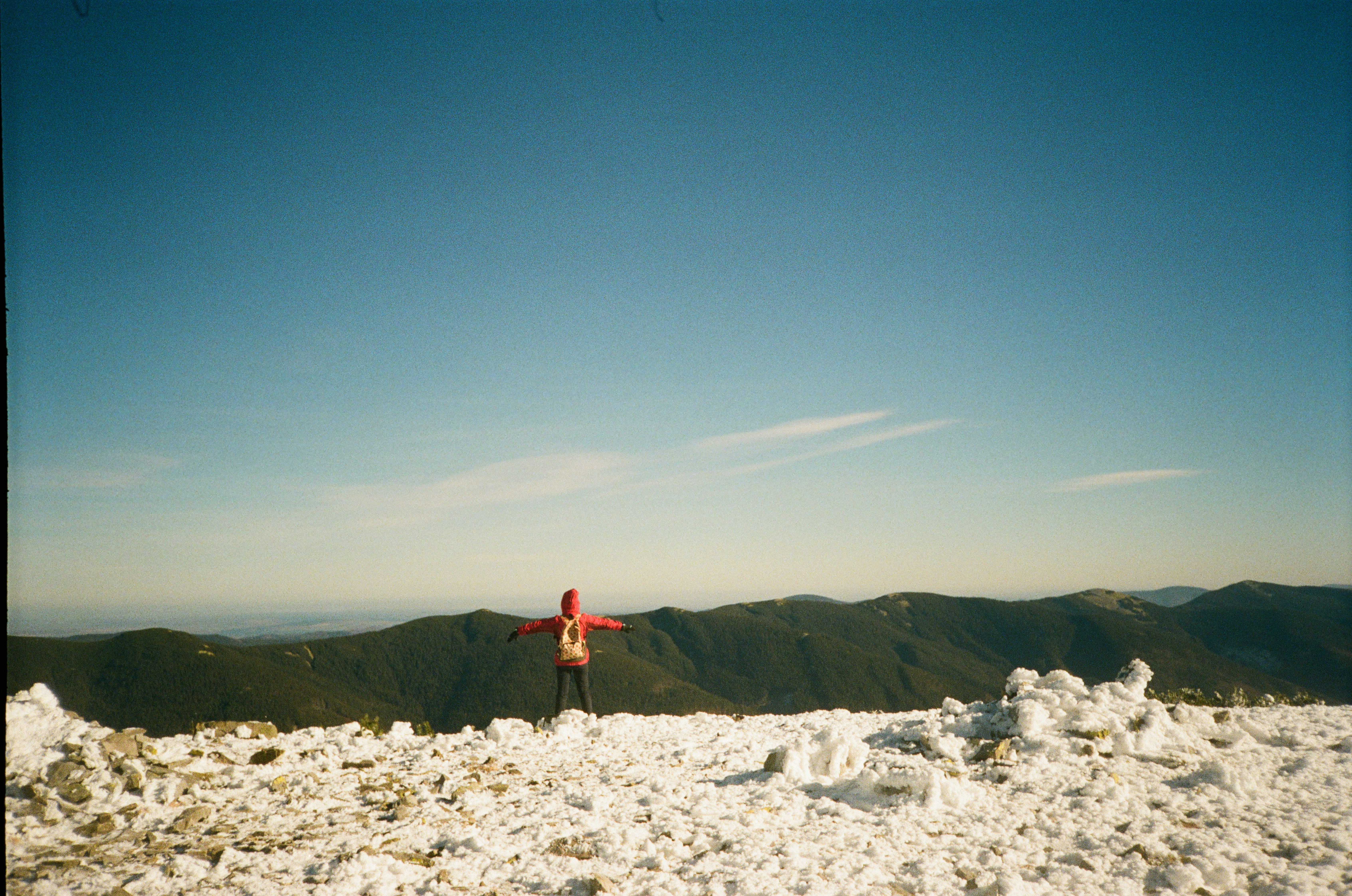 Person in a red jacket stands on snow-covered mountain with arms outstretched, facing distant green hills under a clear blue sky.
