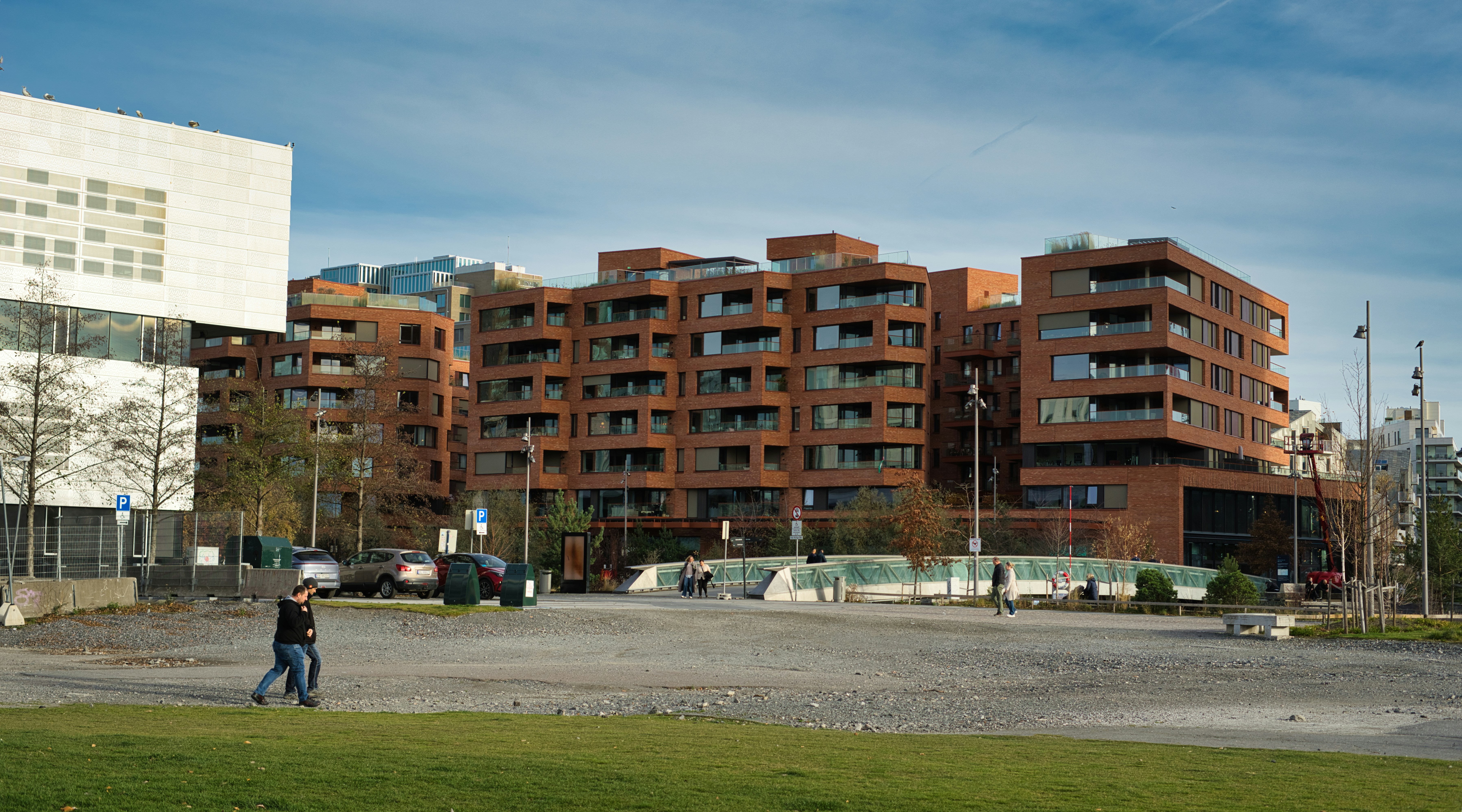 Red brick apartment buildings against a clear sky, with a nearby parking area and pedestrians walking by.