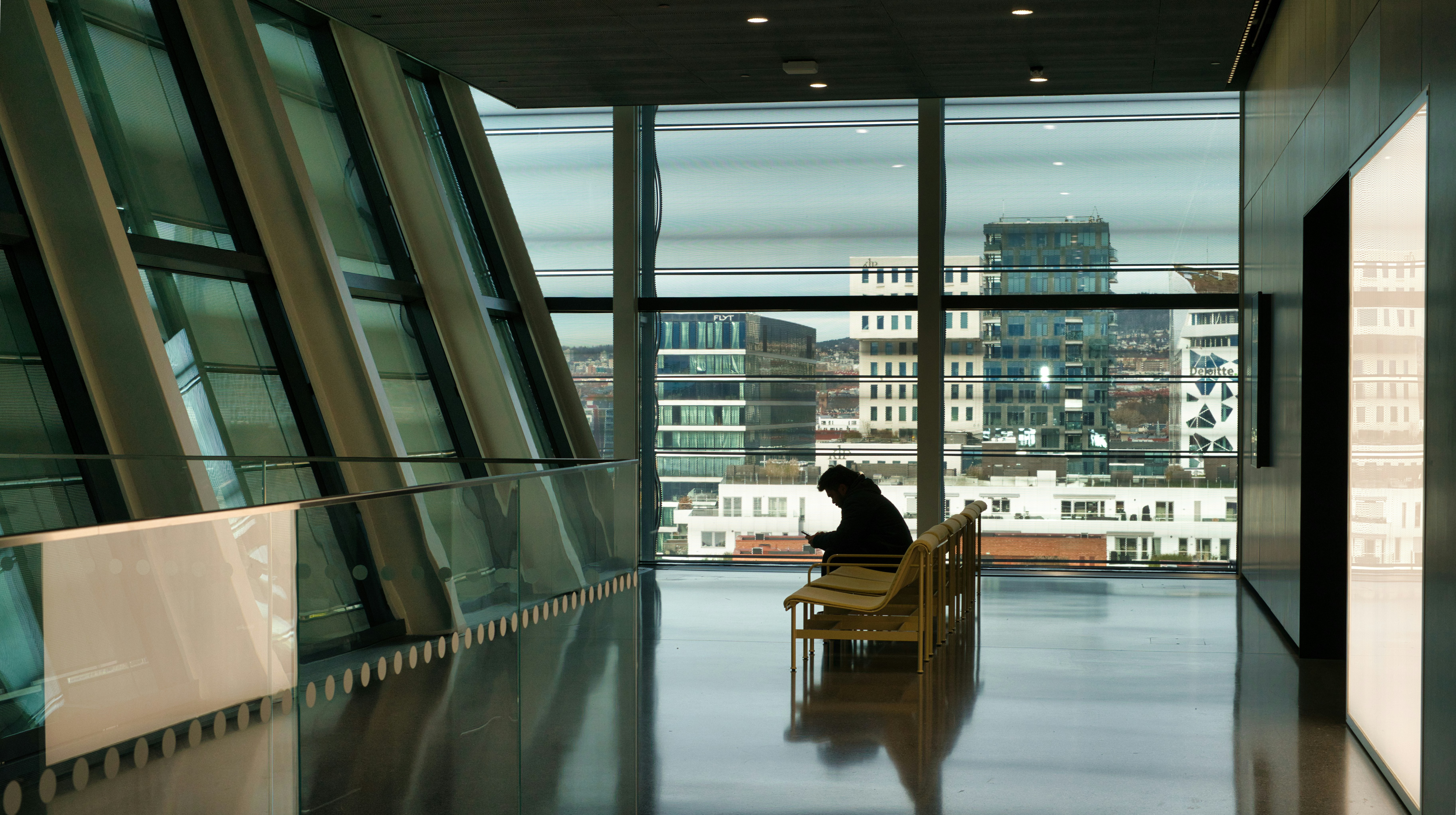 Silhouette of a person seated on a bench in a modern building with large windows framing a cityscape.