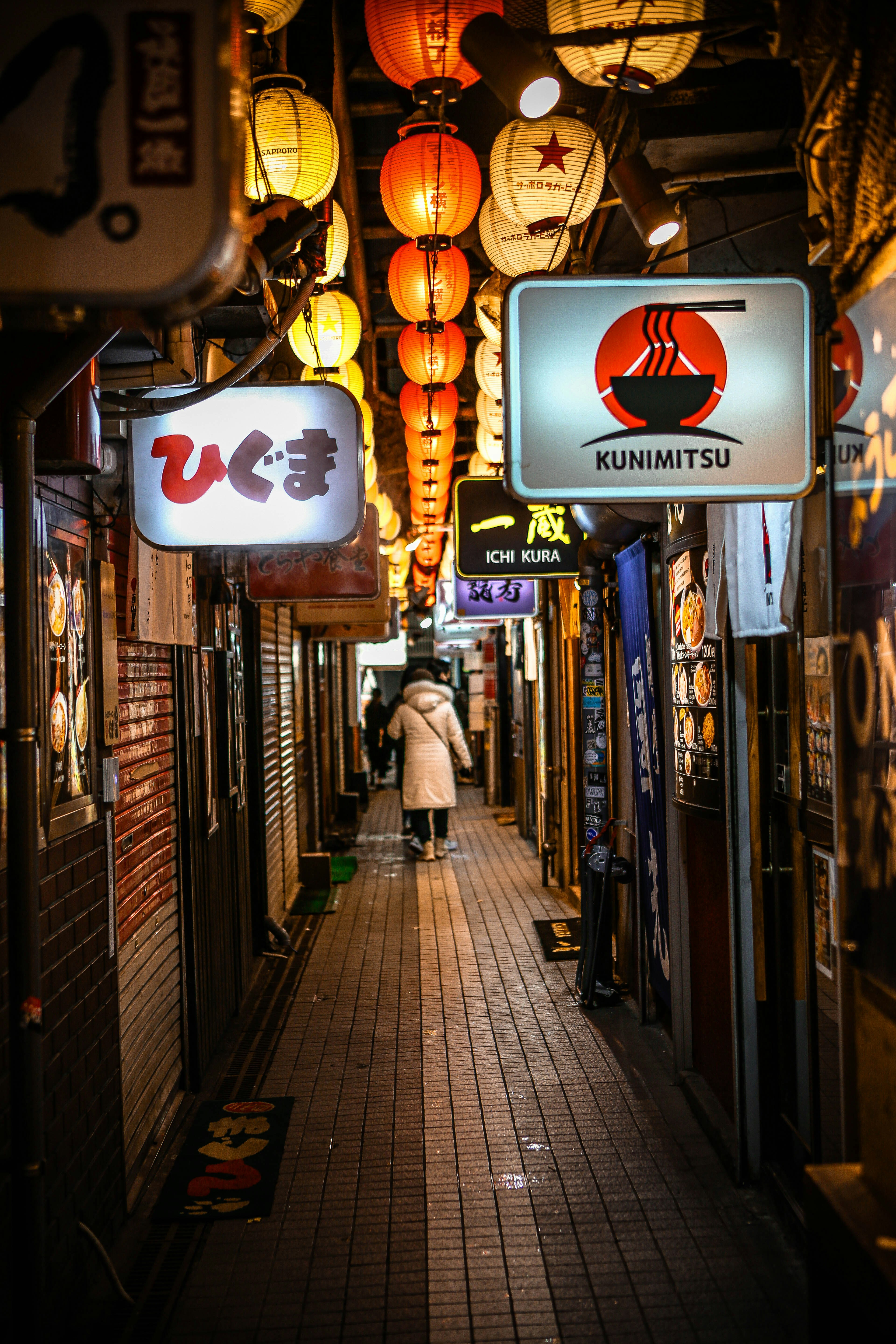 Illuminated alleyway lined with traditional lanterns and restaurant signs, showcasing a vibrant culinary scene. A lone figure strolls through the inviting atmosphere.