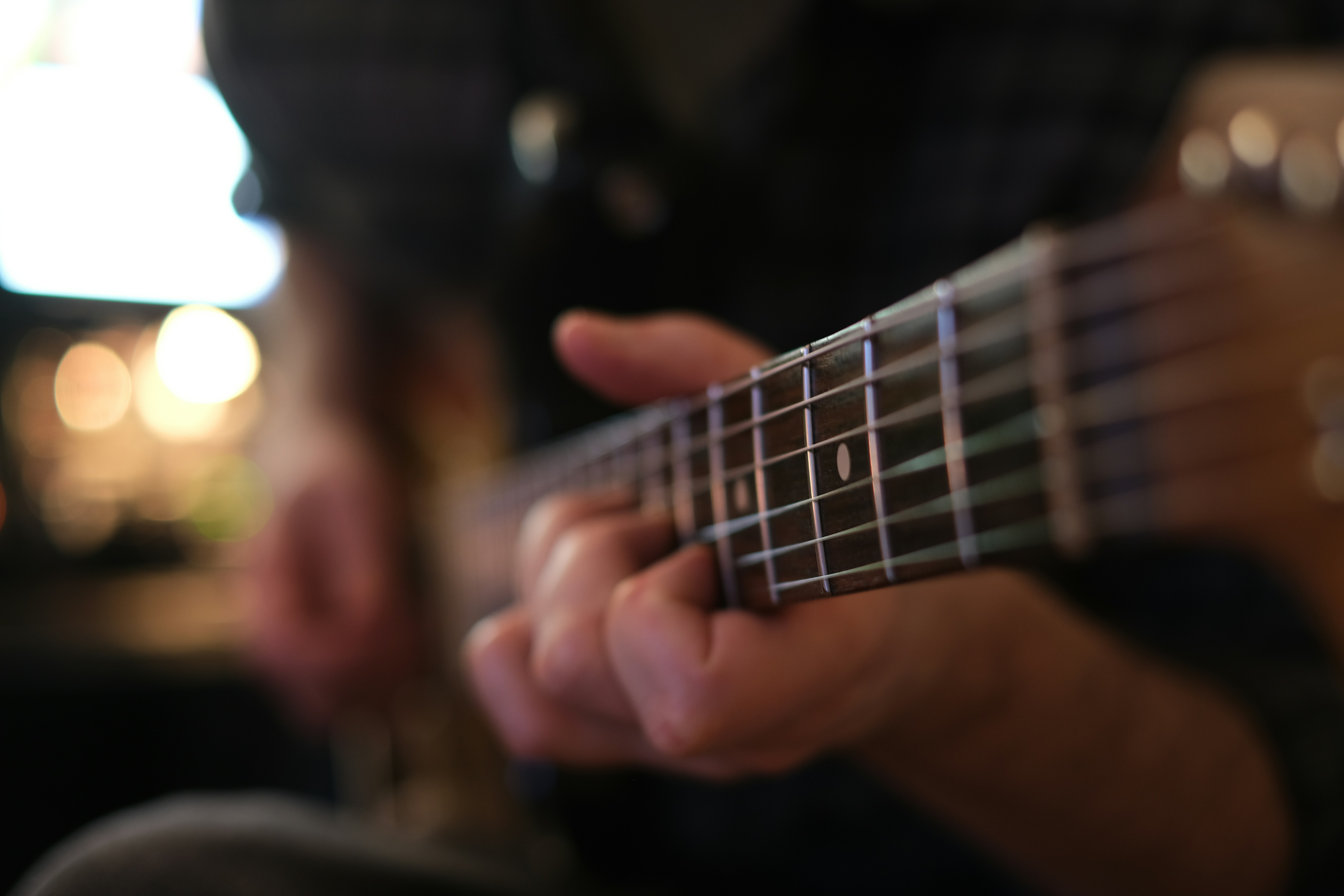 A close-up shot of a guitarist playing an electric guitar, focusing on the fretboard and the player's fingers pressing down on the strings. The shallow depth of field highlights the intricate details of the guitar's neck while softly blurring the background, which appears to be a home studio or practice space with warm ambient lighting. The image captures the essence of musical expression and live performance. | A hand plays guitar, close up.