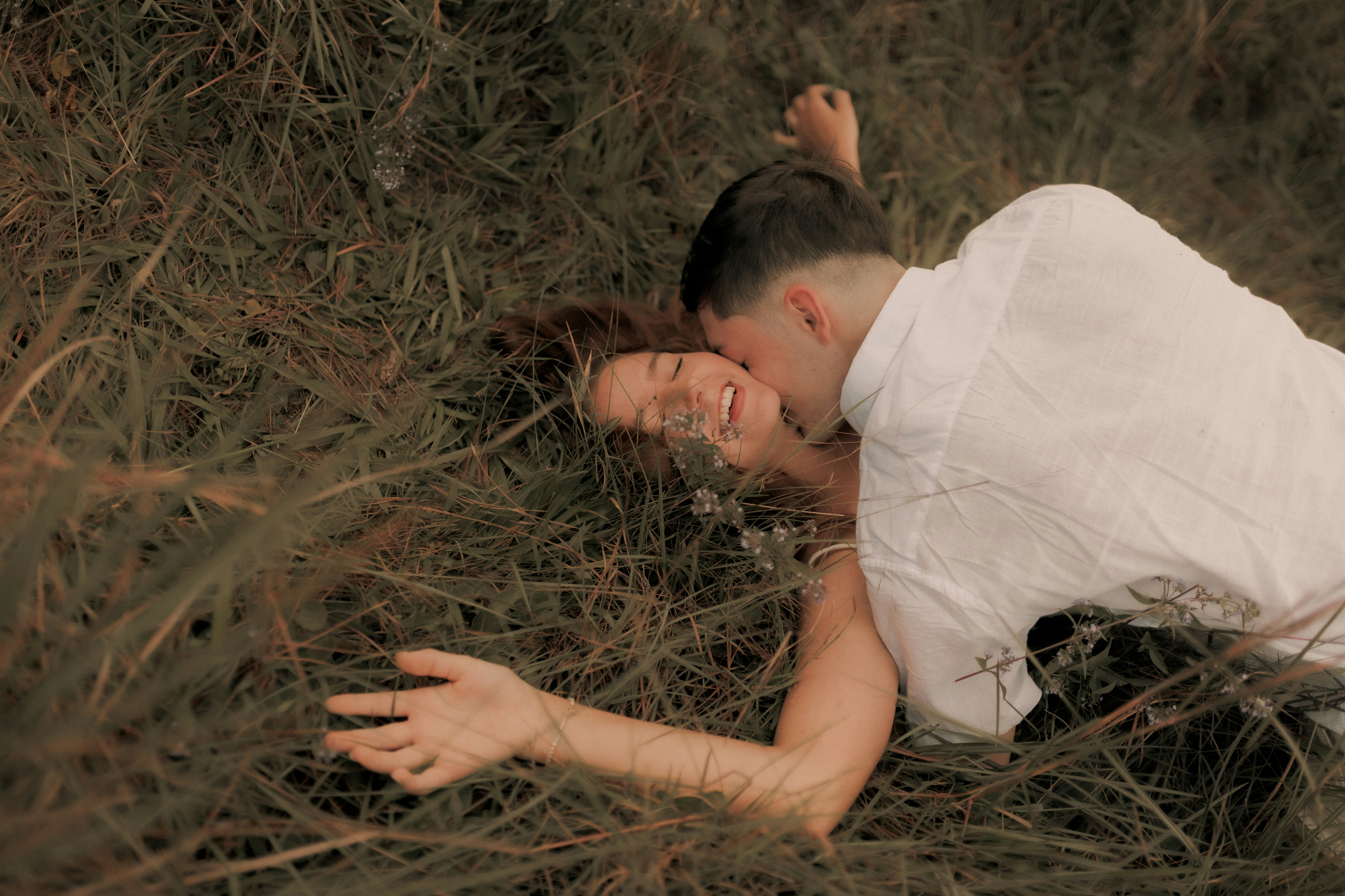Couple lying in tall grass, sharing a tender moment under soft natural light.