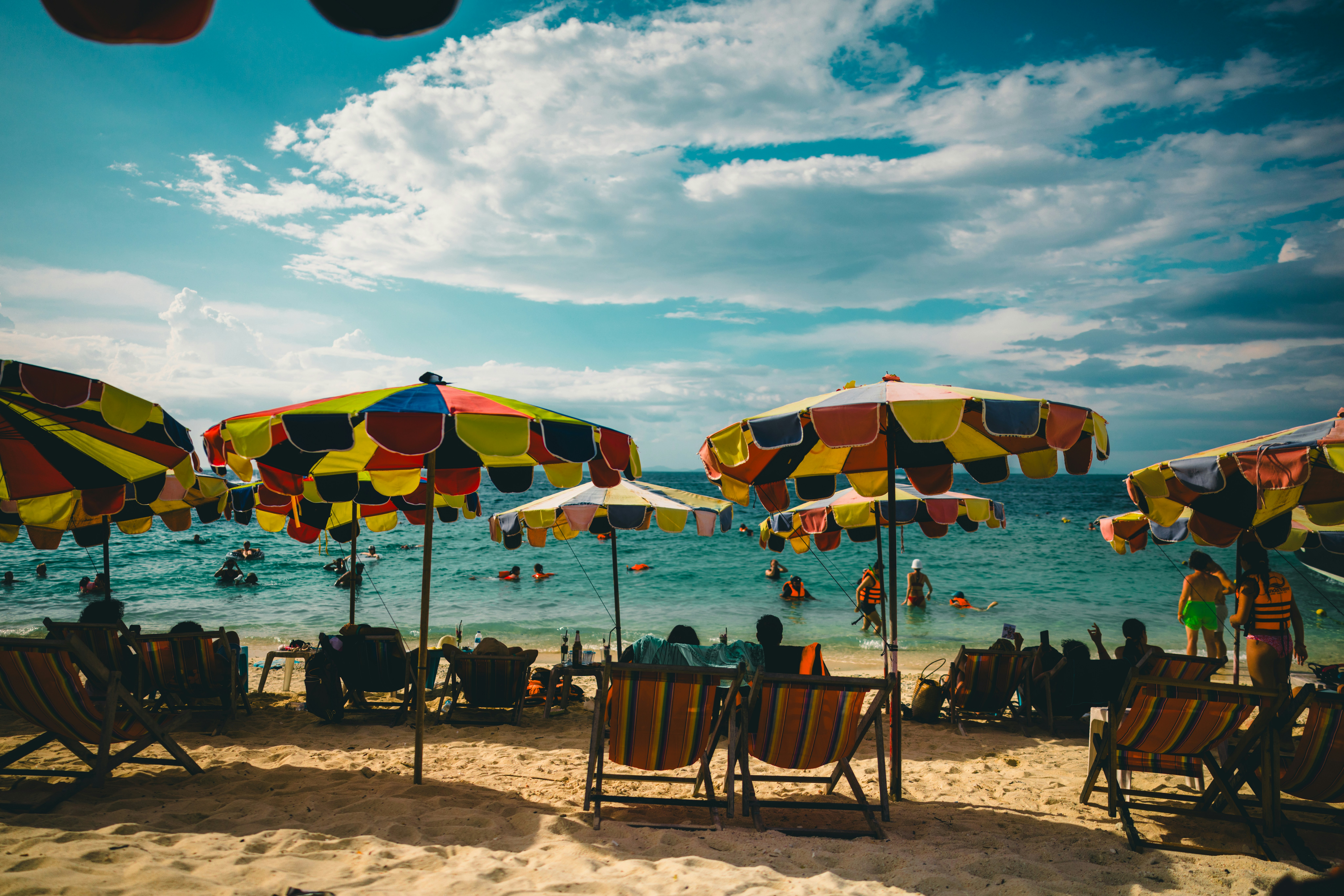 Beachgoers relax under vibrant umbrellas on a sunny shoreline with swimmers in the clear blue sea.