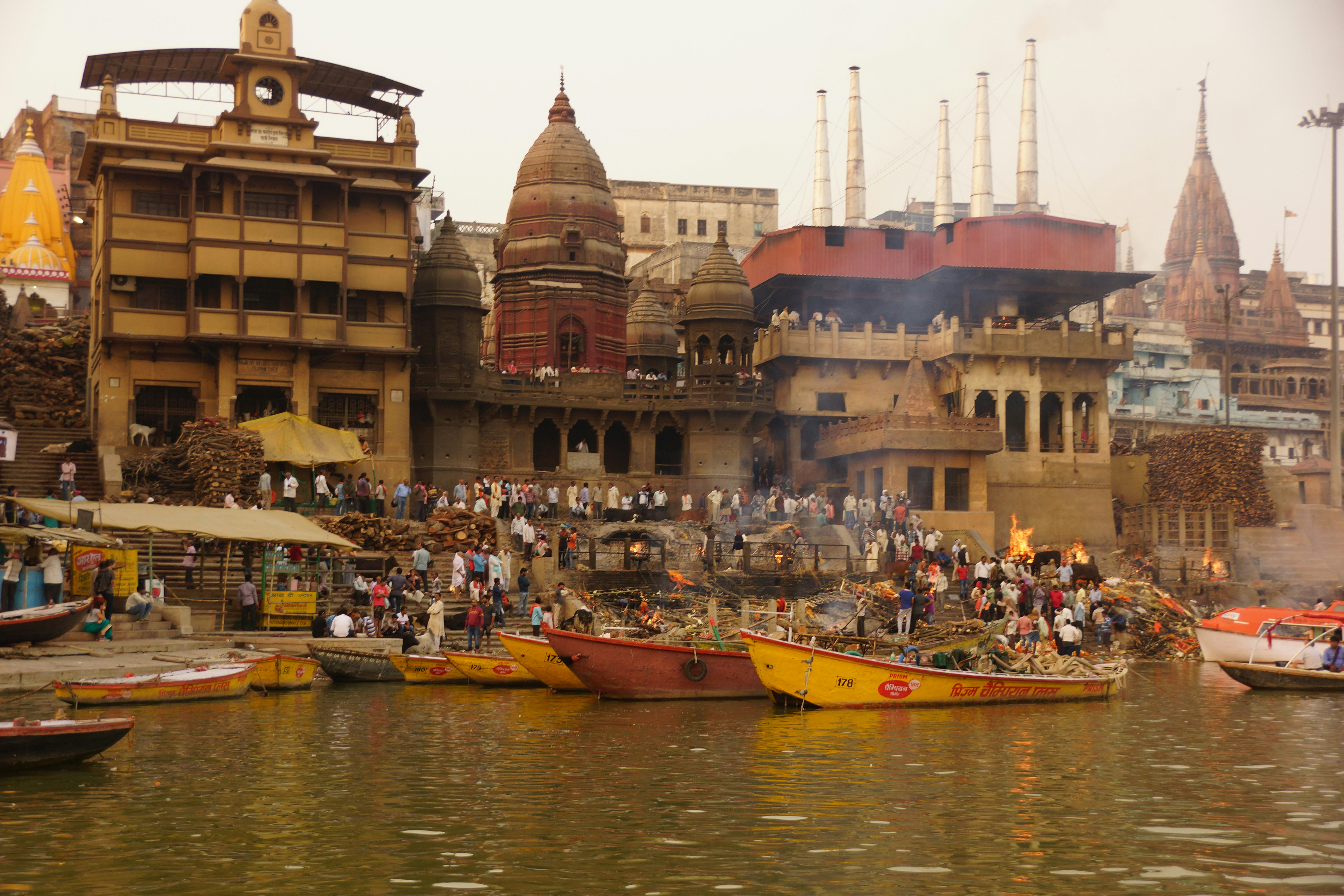 Crowds gather along the Ganges River in Varanasi, with historic architecture and boats lining the waterway.