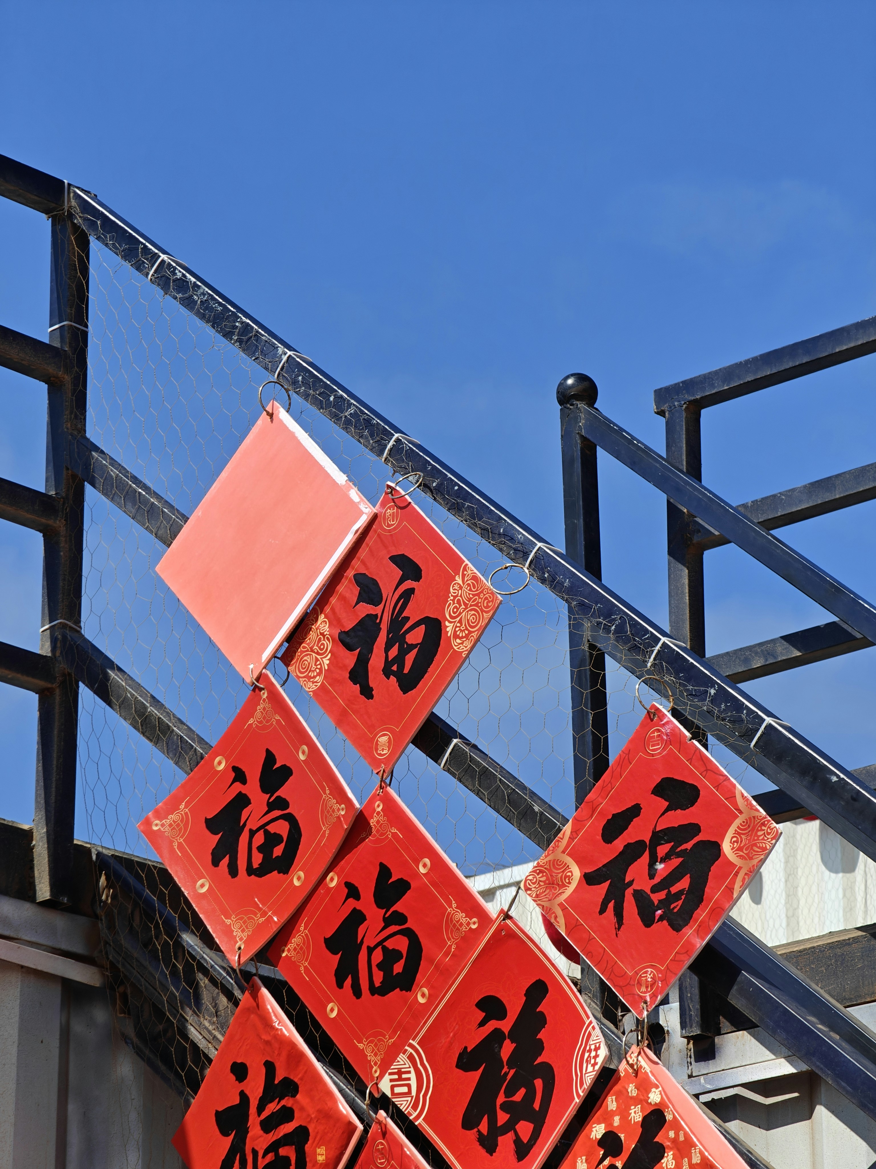 Red Chinese 福 charms hang on a wire-mesh fence along a metal staircase, set against a bright blue sky.