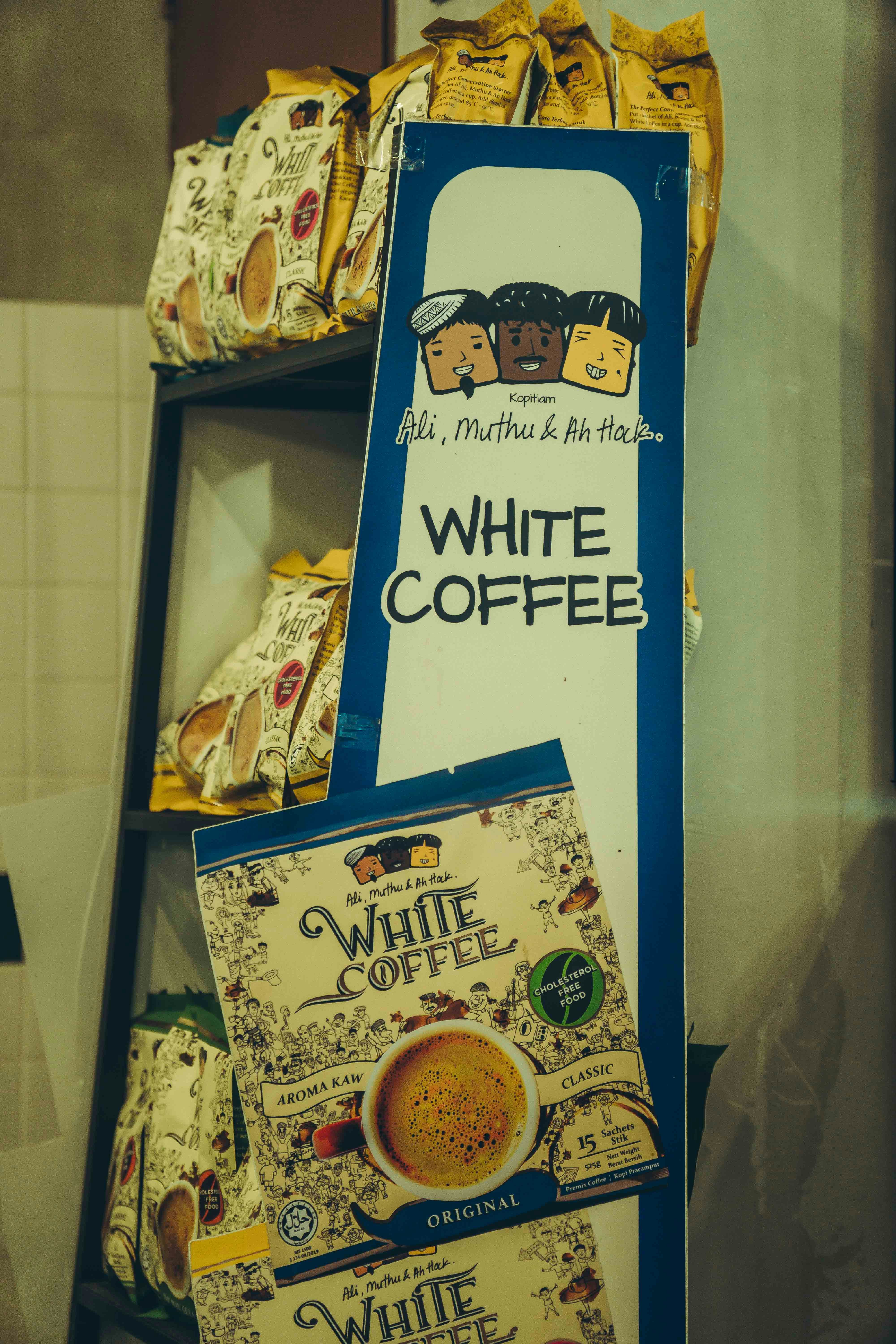 A vibrant display featuring a sign and packaging for White Coffee, showcasing playful illustrations of three characters. The branding emphasizes the product's unique qualities.