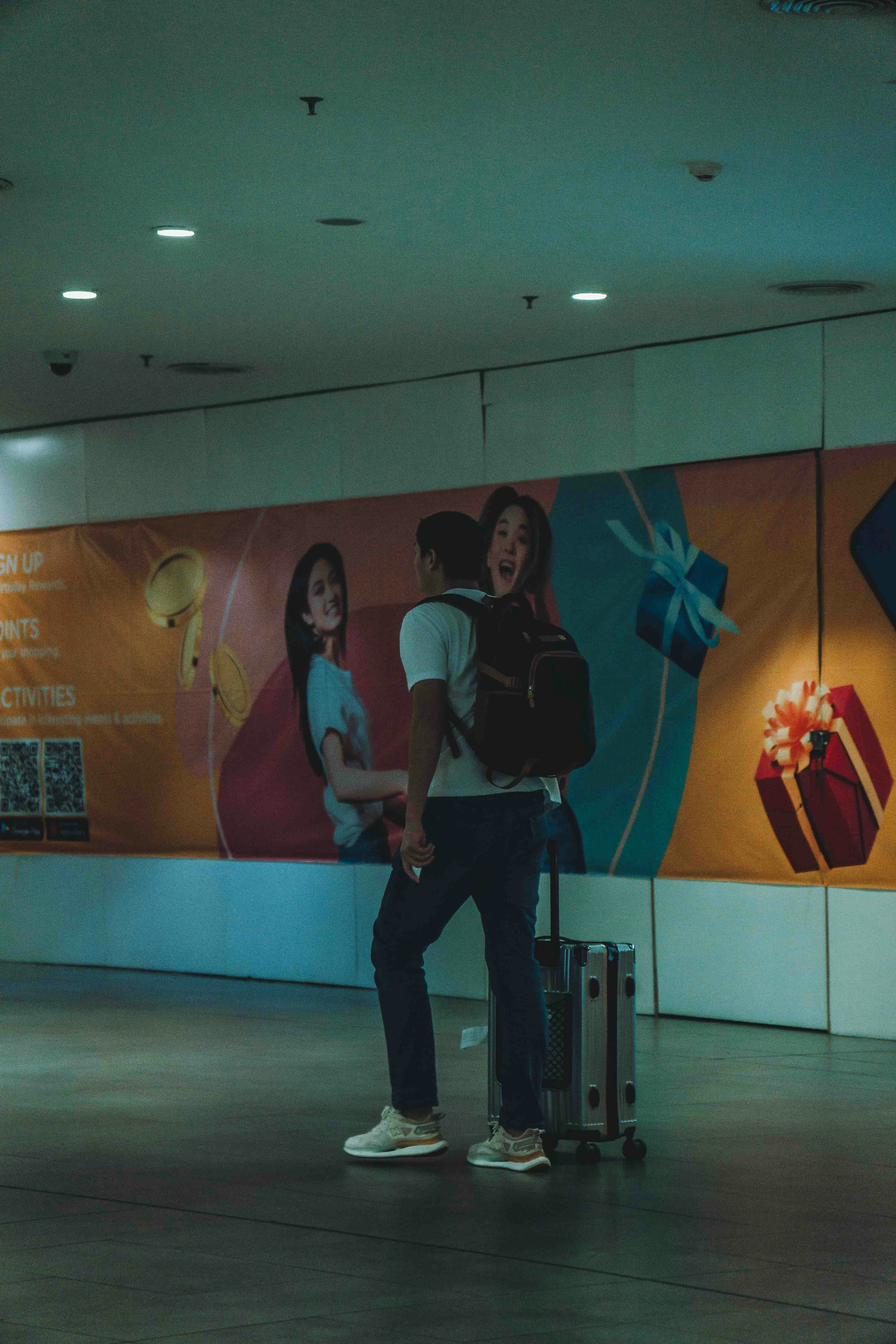 Traveler with suitcase walks past a colorful advertisement in a dimly lit corridor.