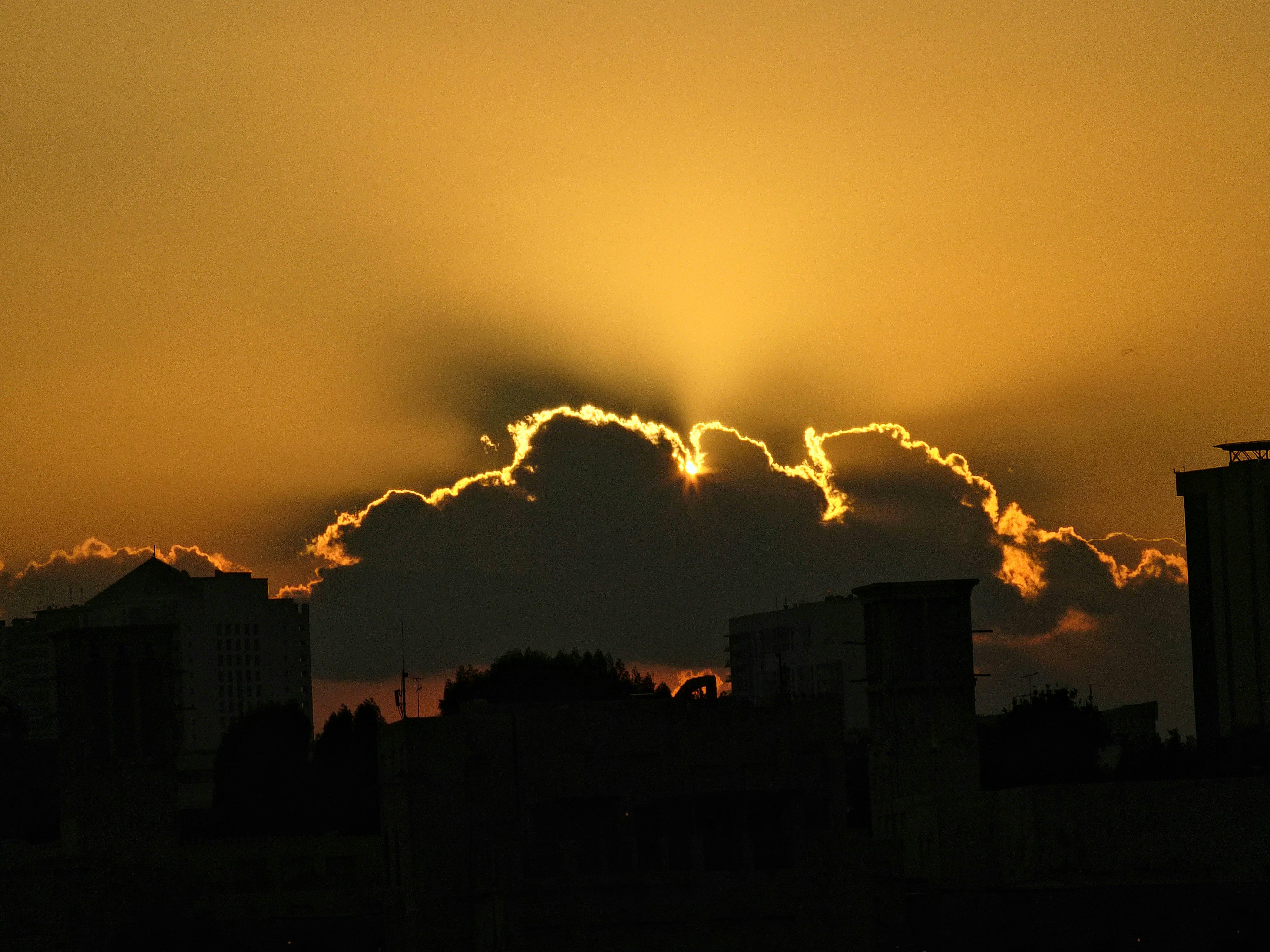 Sun peeking over city skyline, casting a golden glow on silhouetted buildings and clouds.