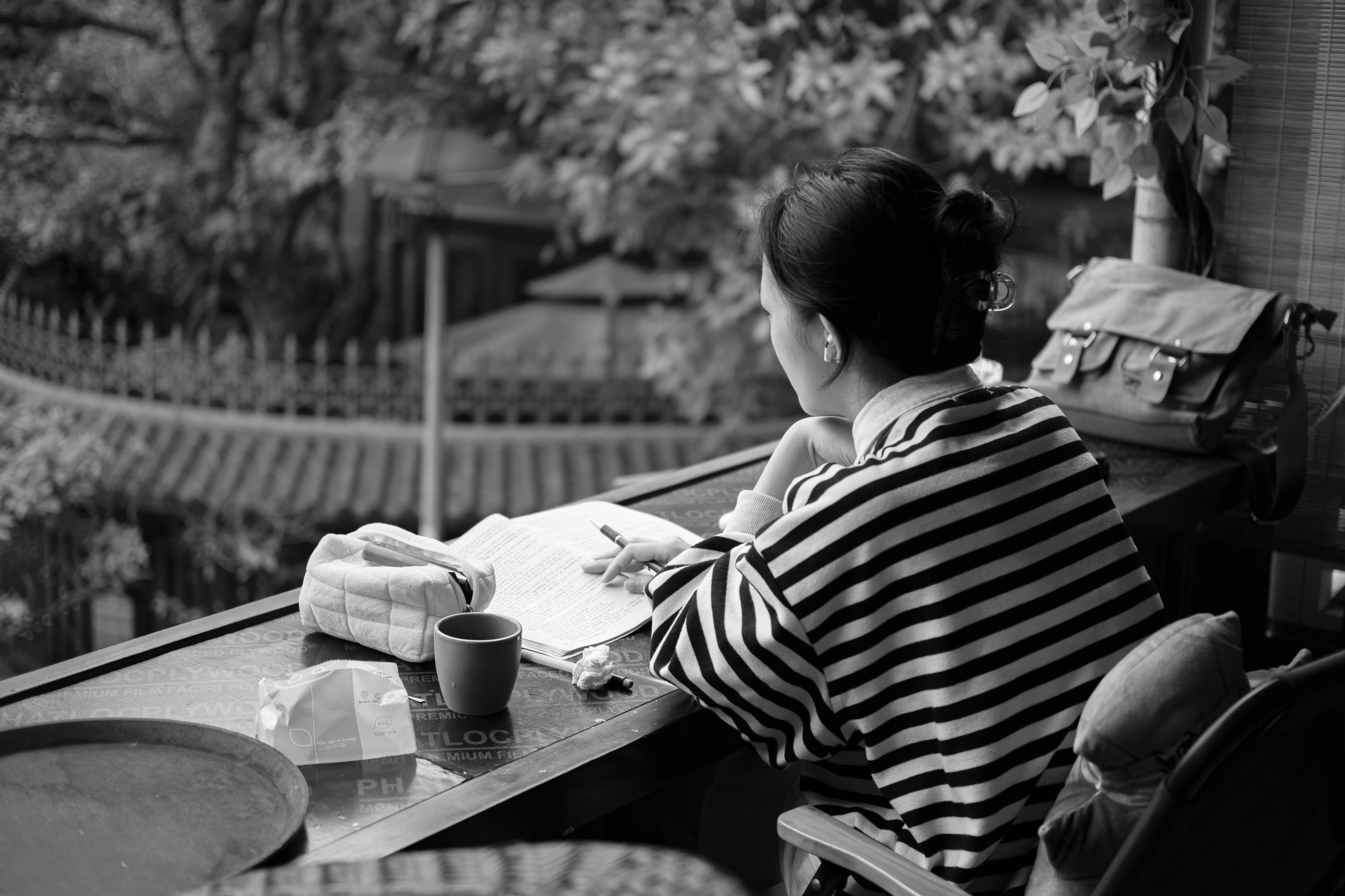 Person in striped shirt writing at a window-side desk with trees outside.