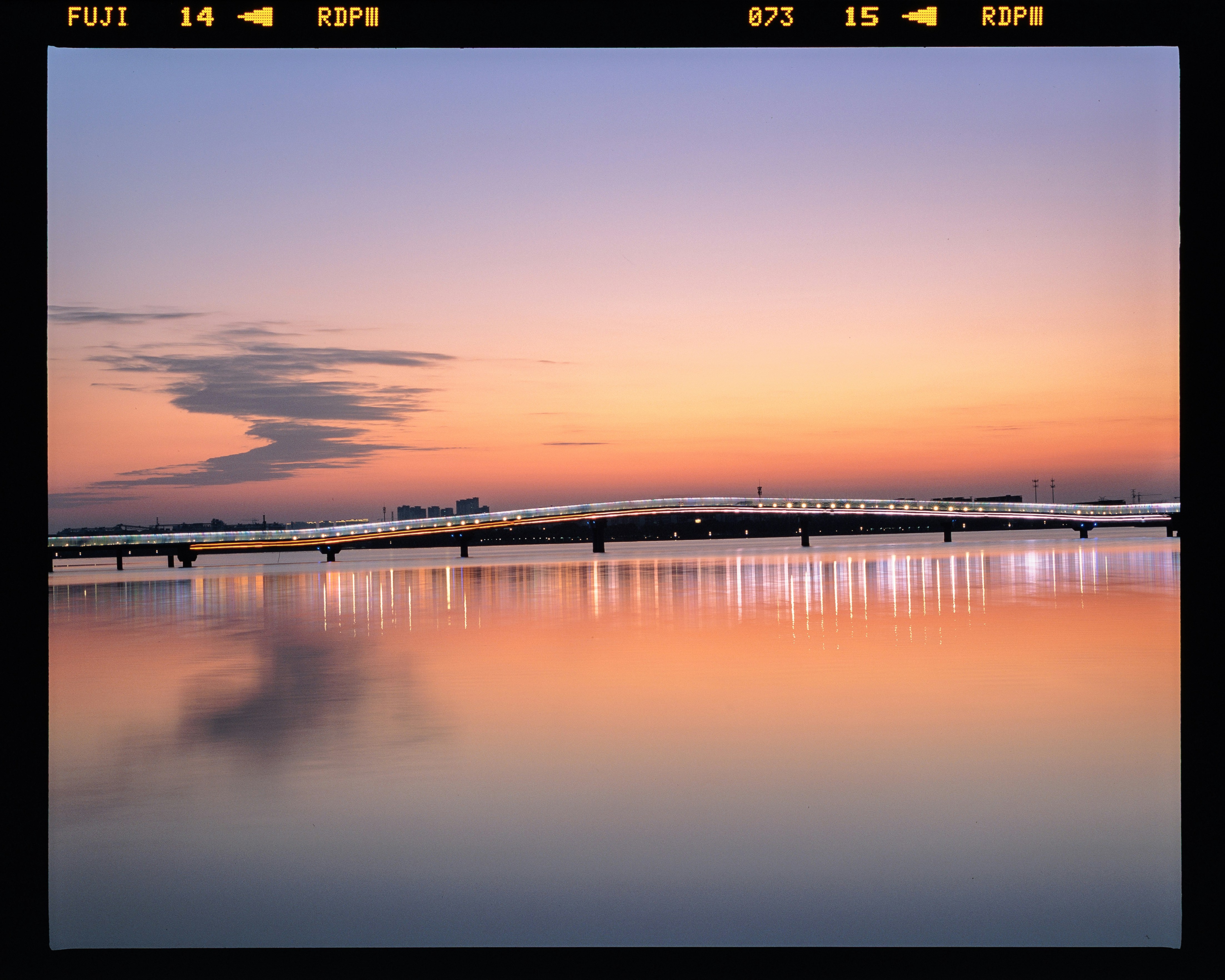 Serene sunset sky reflecting on a calm river with a lit bridge stretching across the horizon.