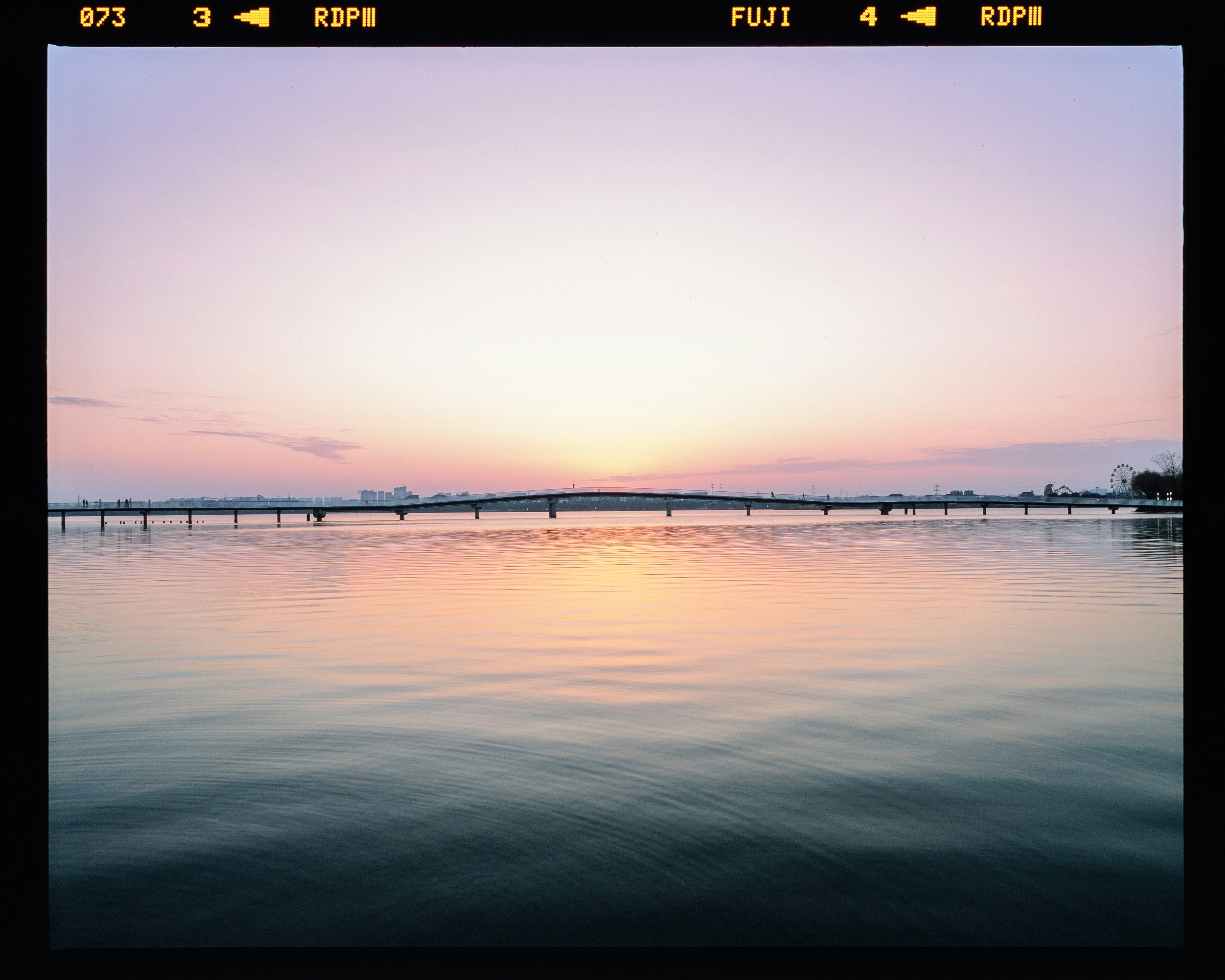 Sunrise over a calm lake with a silhouetted bridge and soft pastel sky reflections.