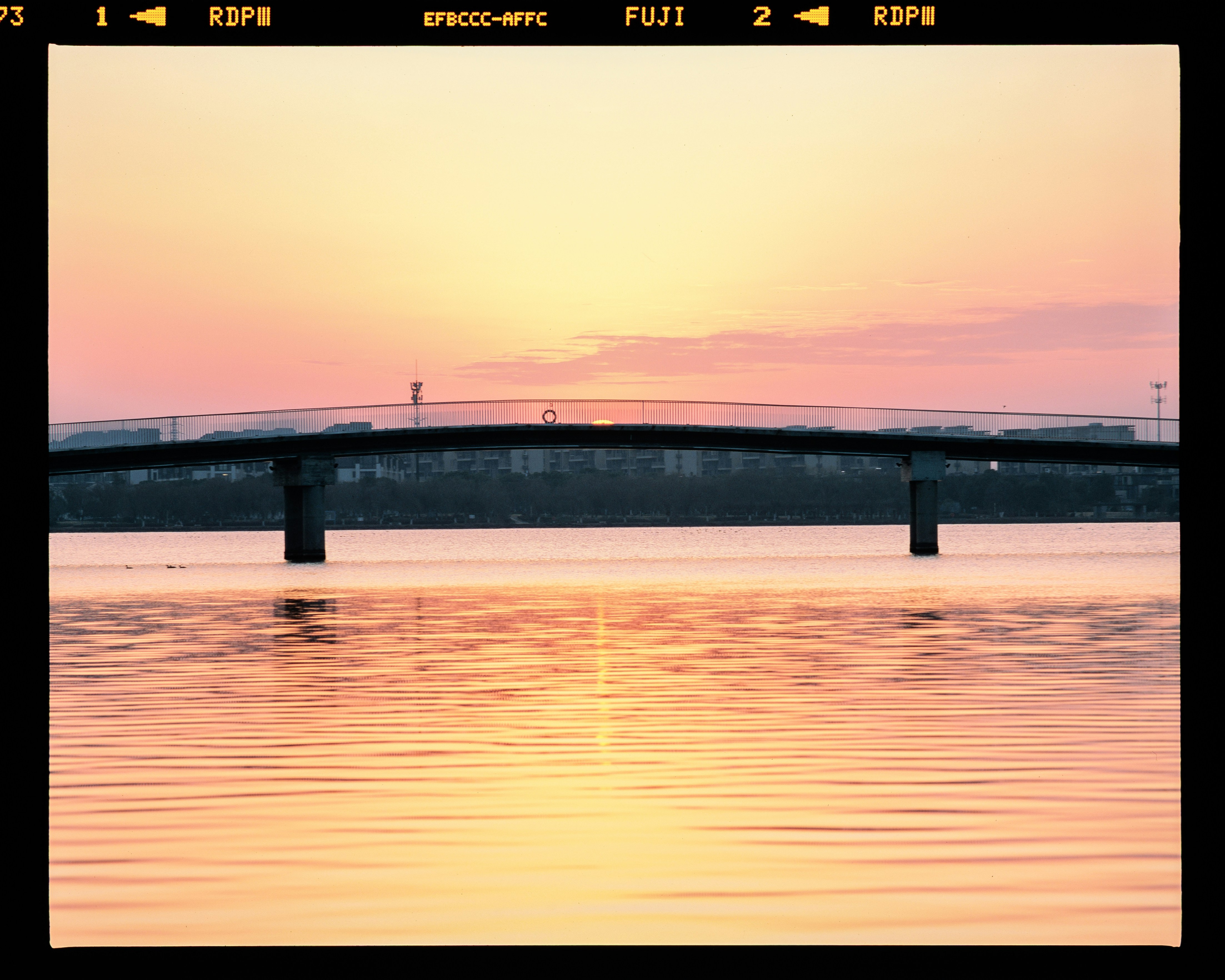 Sunset hues reflect on calm water beneath a silhouetted bridge, with a city skyline in the distance.