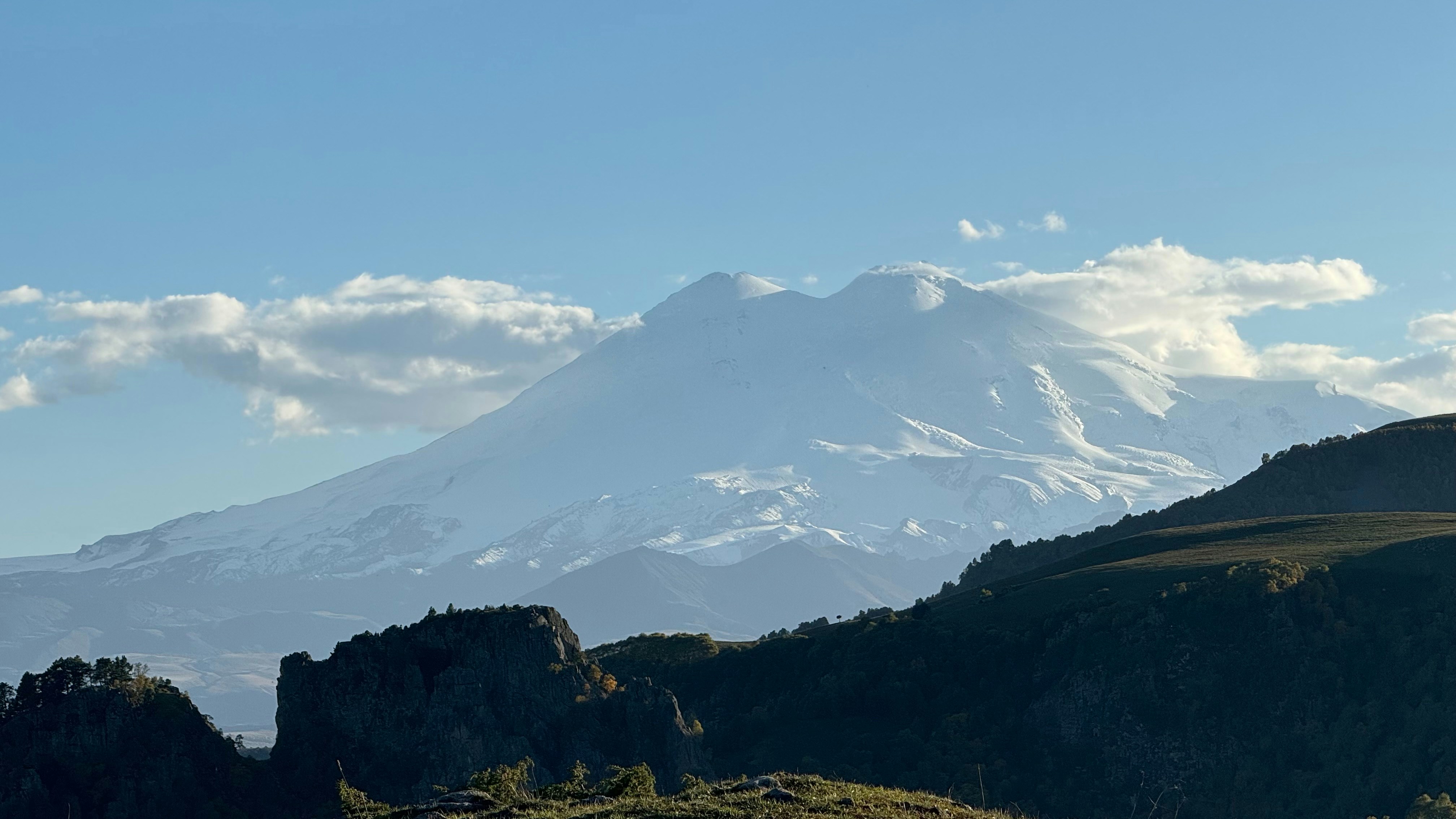 Snow-covered mountain peaks rise beneath scattered clouds in a clear blue sky.