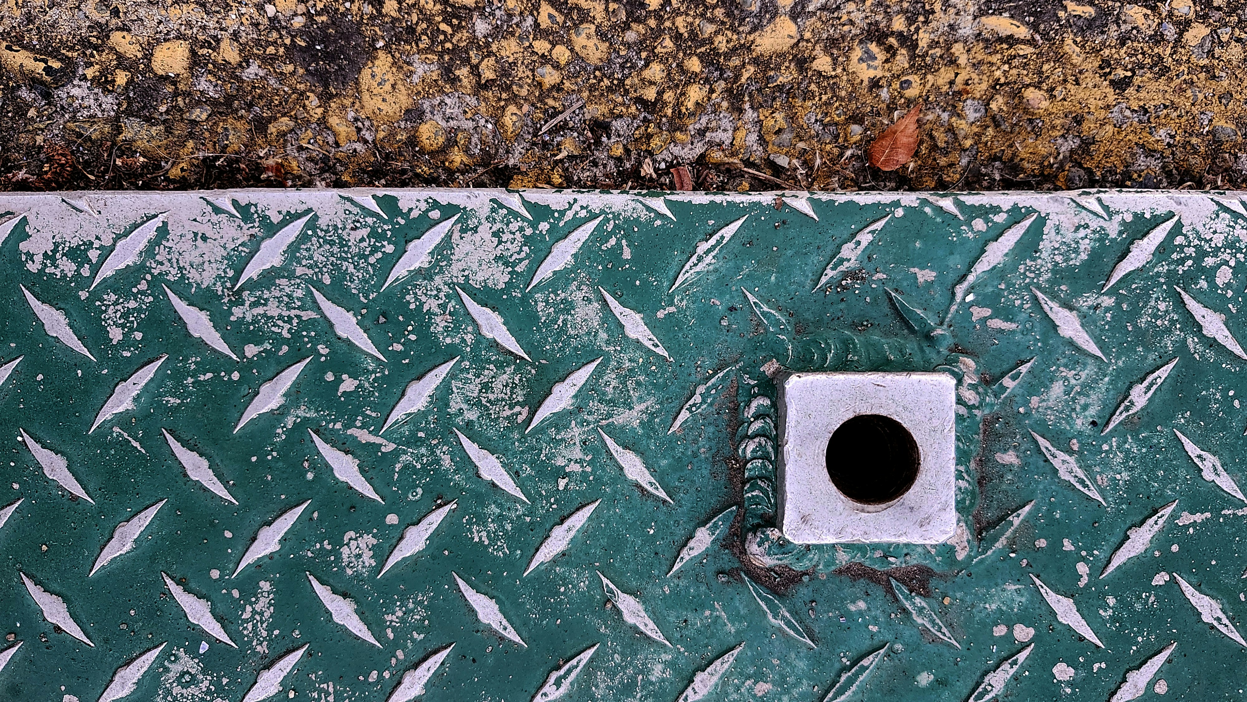 Close-up of a green diamond-patterned metal surface with a square hole, juxtaposed against a textured dirt background. The image highlights contrasting materials and urban elements.
