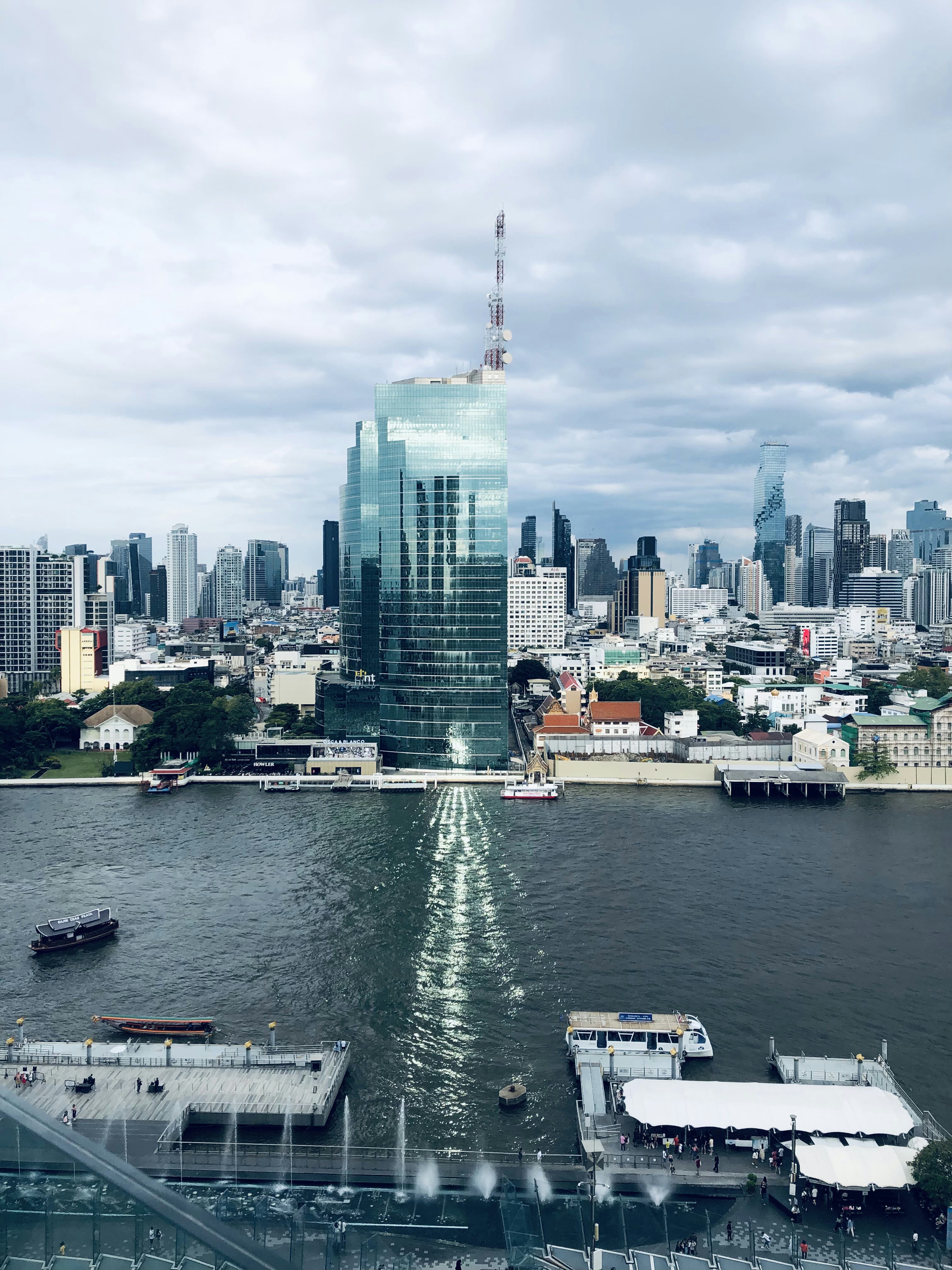 Gleaming glass skyscraper reflects sunlight onto the Chao Phraya River as boats glide through the water under a cloudy sky.
