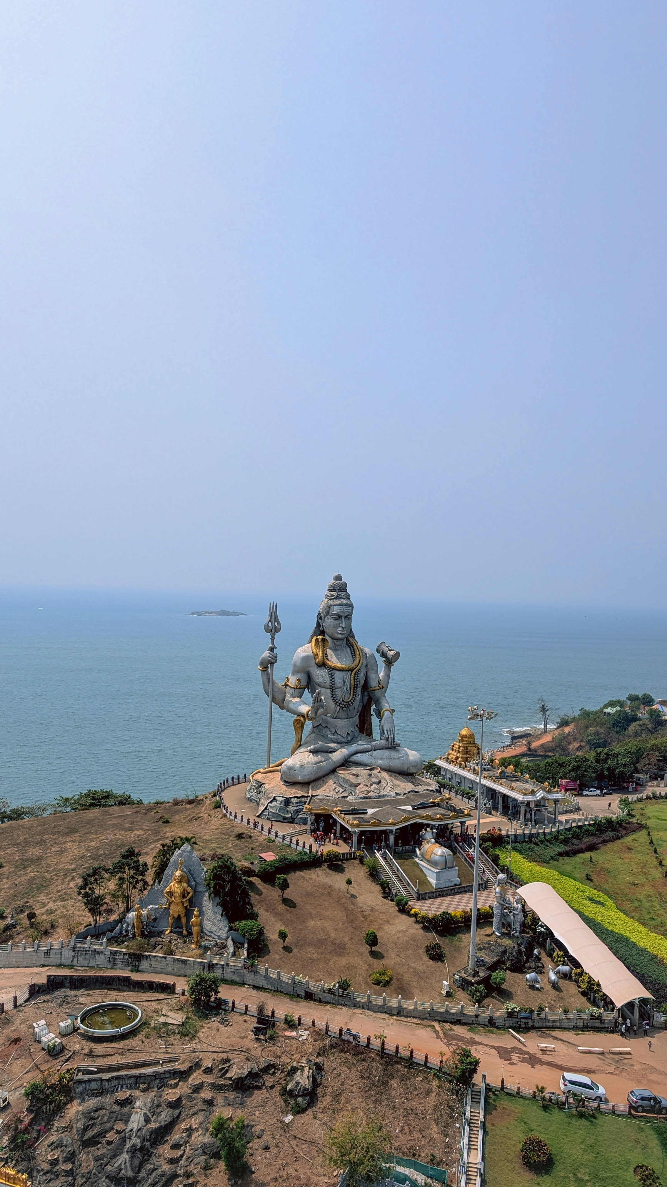 Colossal seated deity atop a hillside temple complex overlooks the sea, with winding paths and smaller statues spread across the grounds toward the horizon.