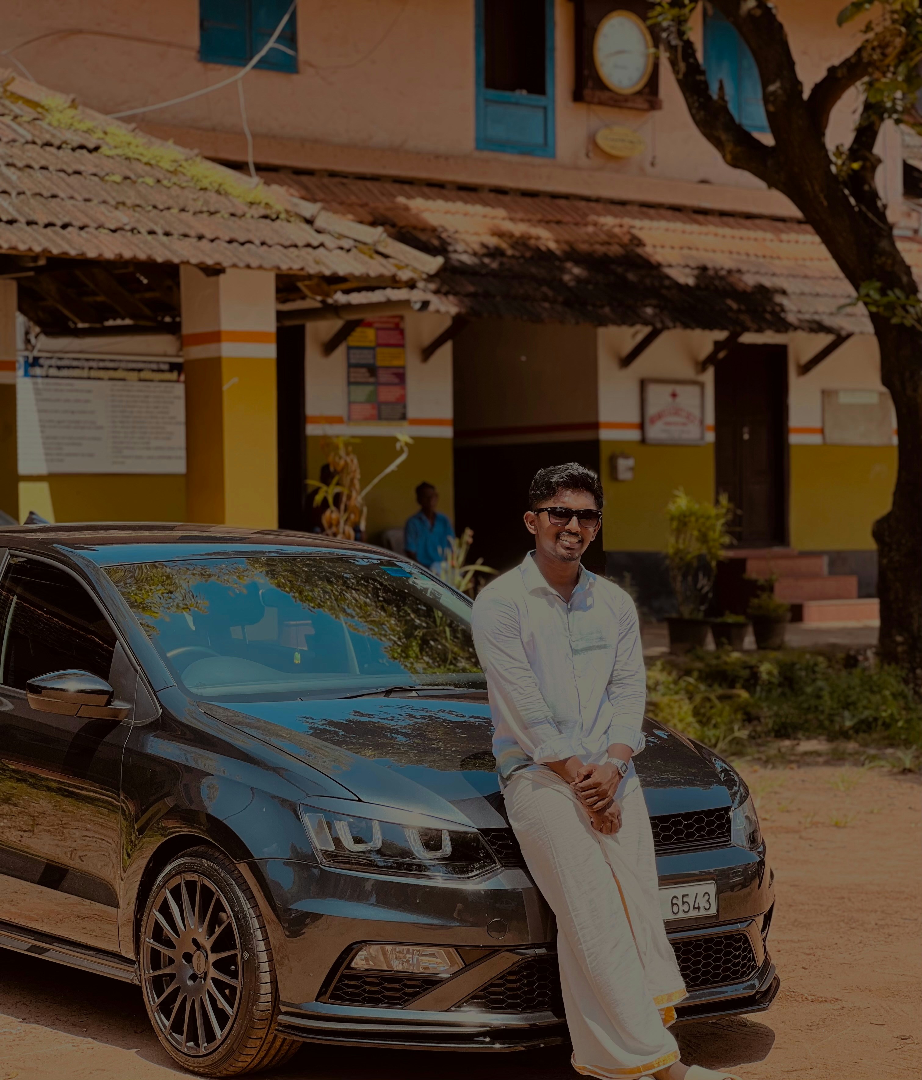 Man in white shirt leaning on a dark sedan in a sunlit campus courtyard