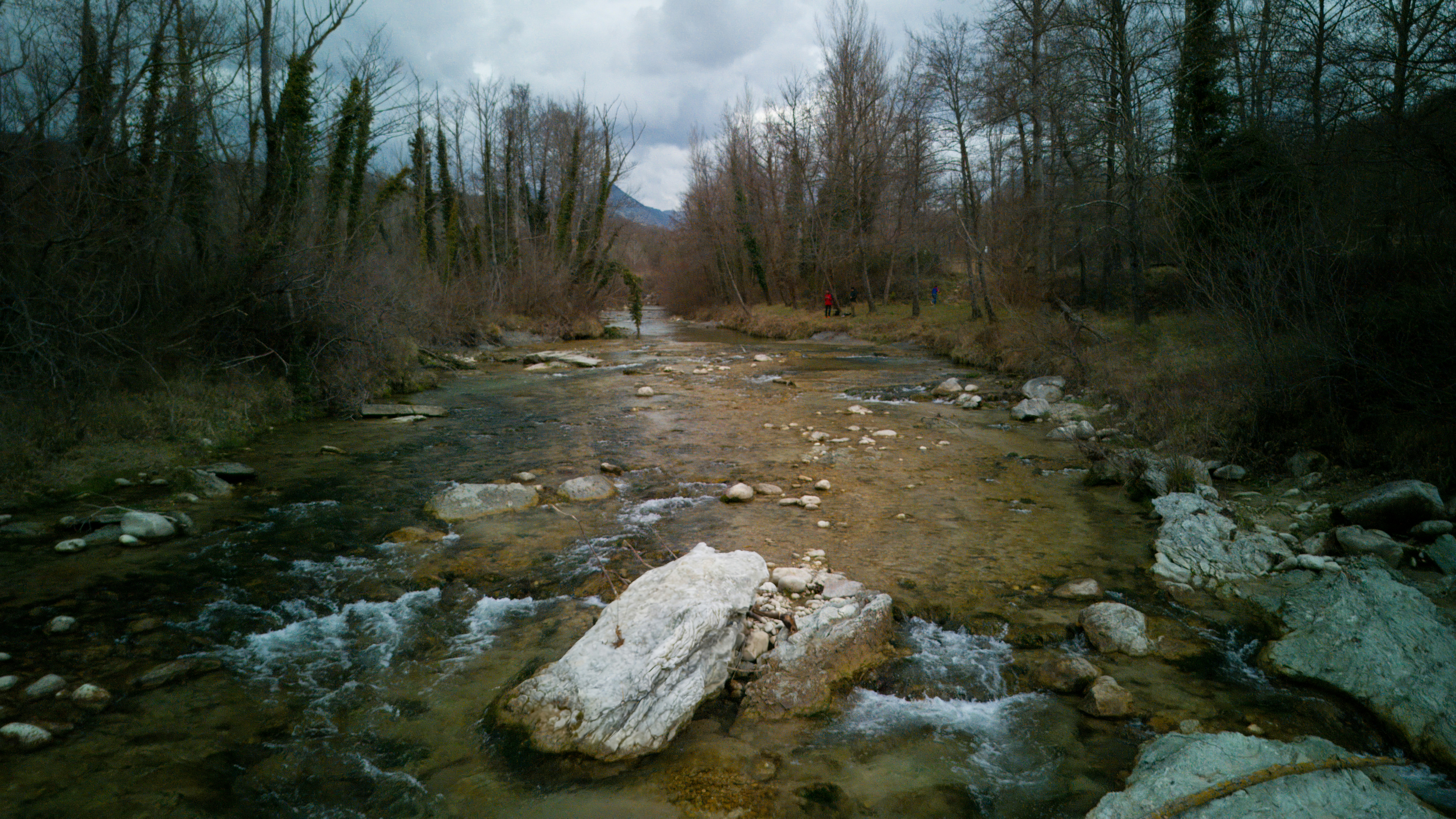 A serene river meanders through a landscape of bare trees and smooth stones, reflecting the overcast sky. The scene evokes a sense of calm and connection to nature.