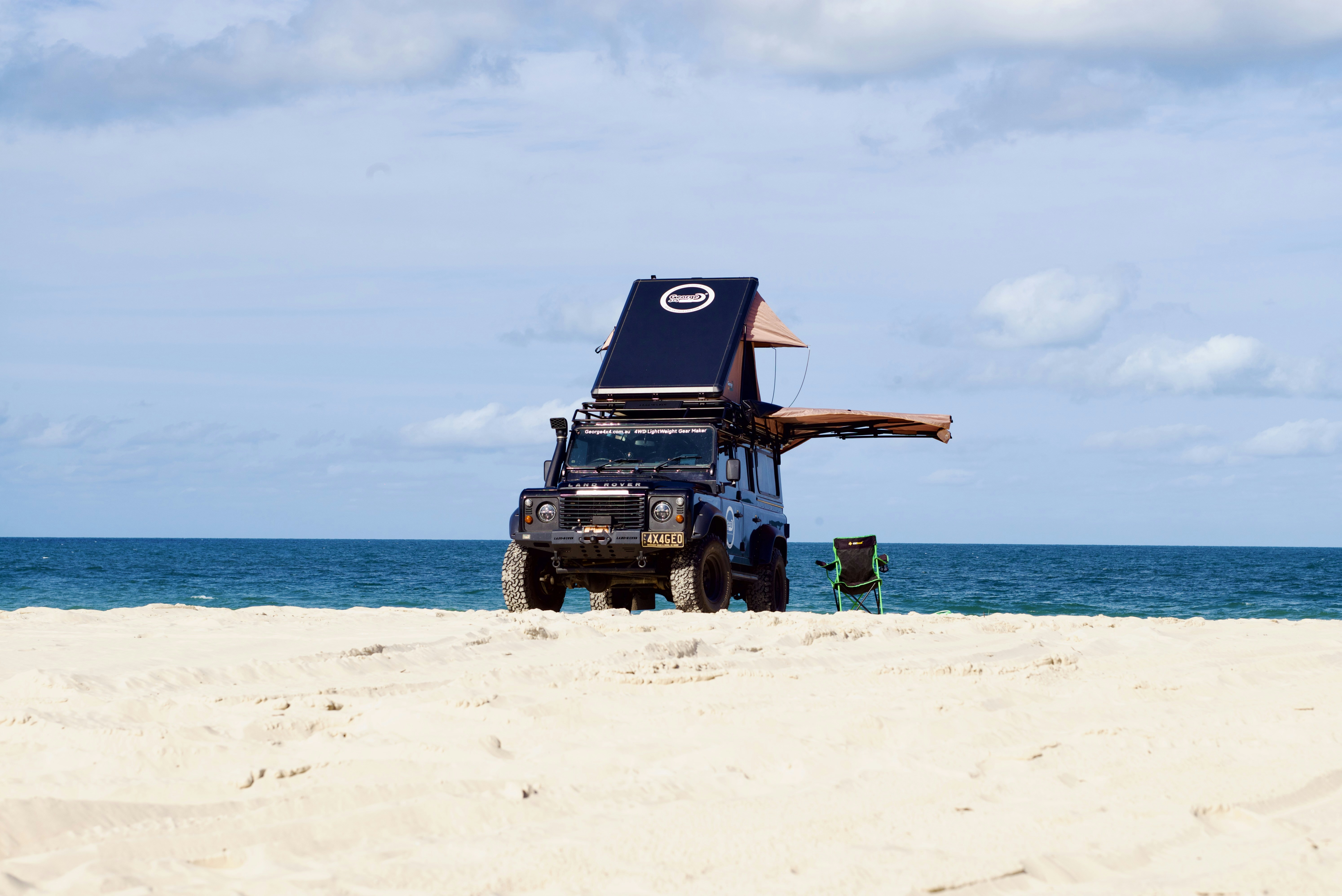 Off-road vehicle with a rooftop tent set up on a sandy beach near the ocean under a clear blue sky.