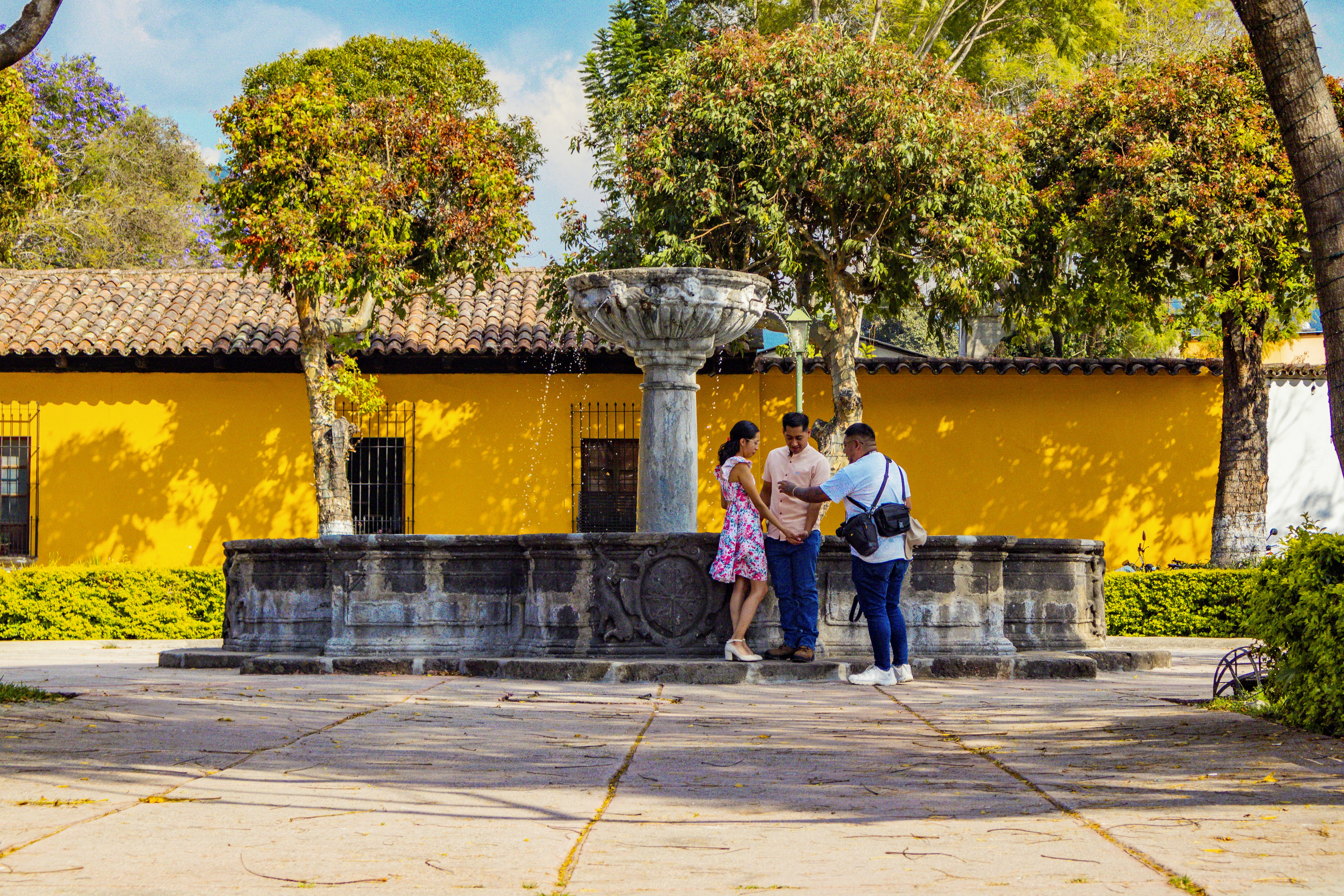 Group of people gather around a stone fountain in a vibrant park with a yellow wall backdrop.