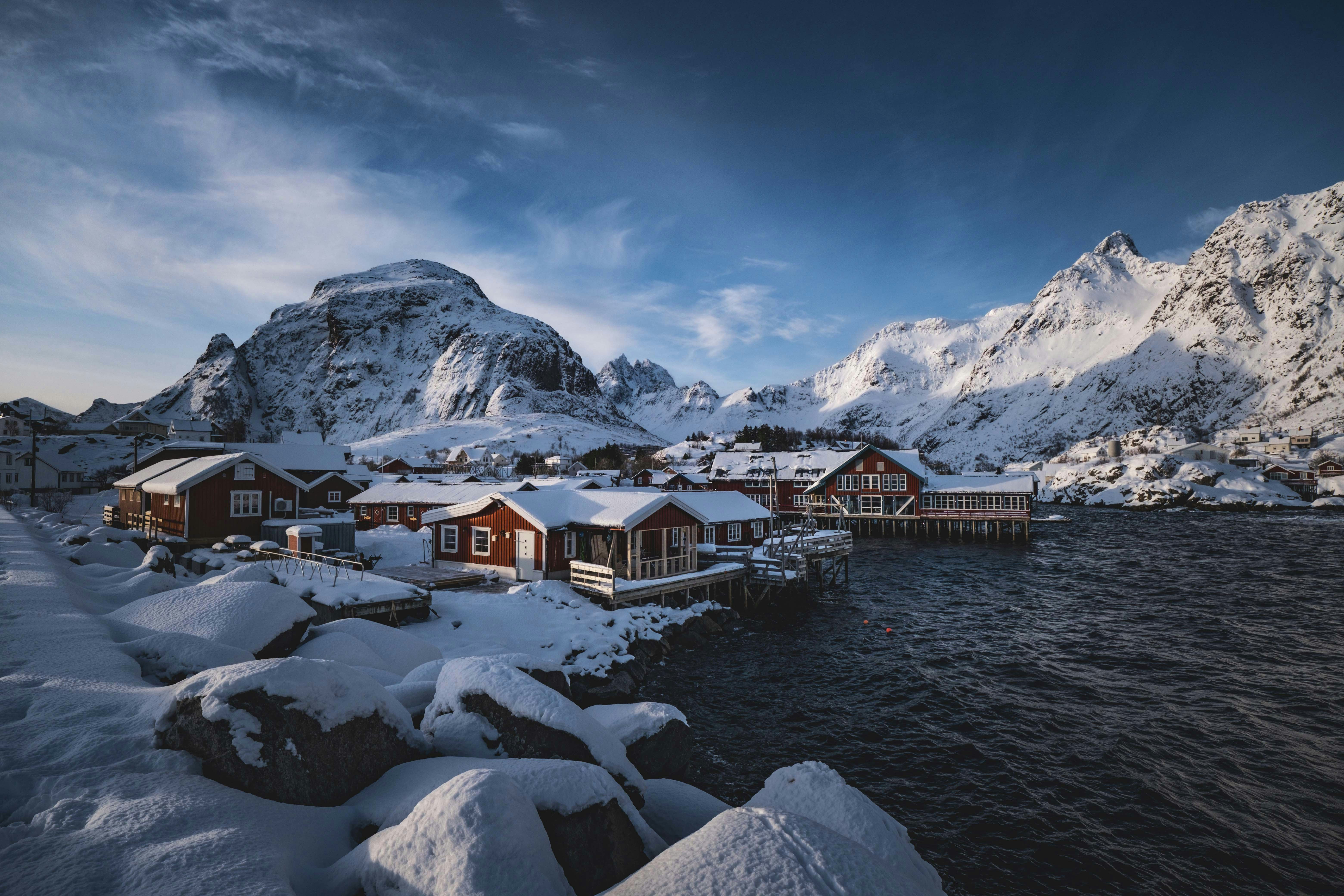 Red cabins nestled against snowy mountains by a dark blue fjord.