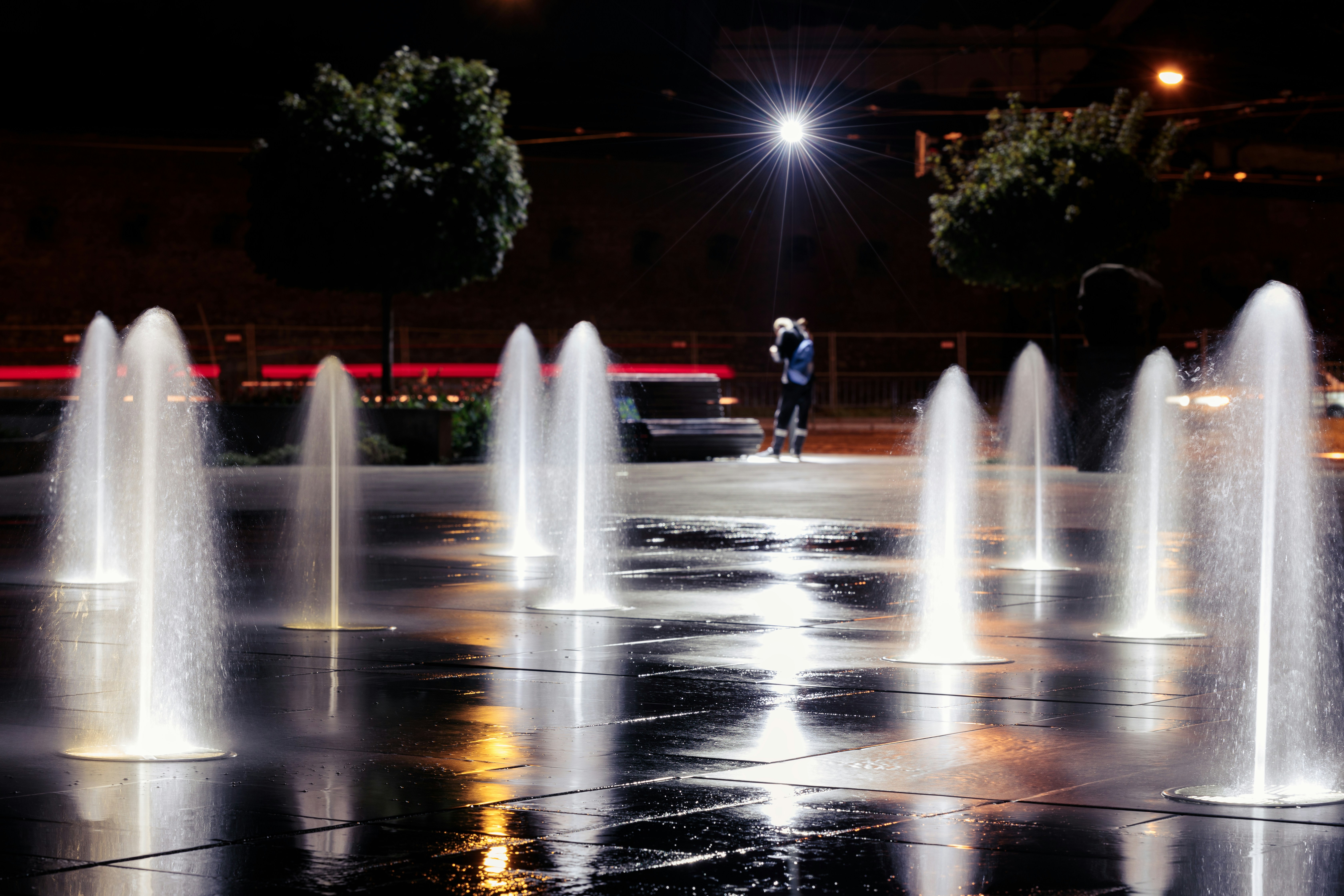 Illuminated fountains dance in a city square at night with a blurred figure in the background.