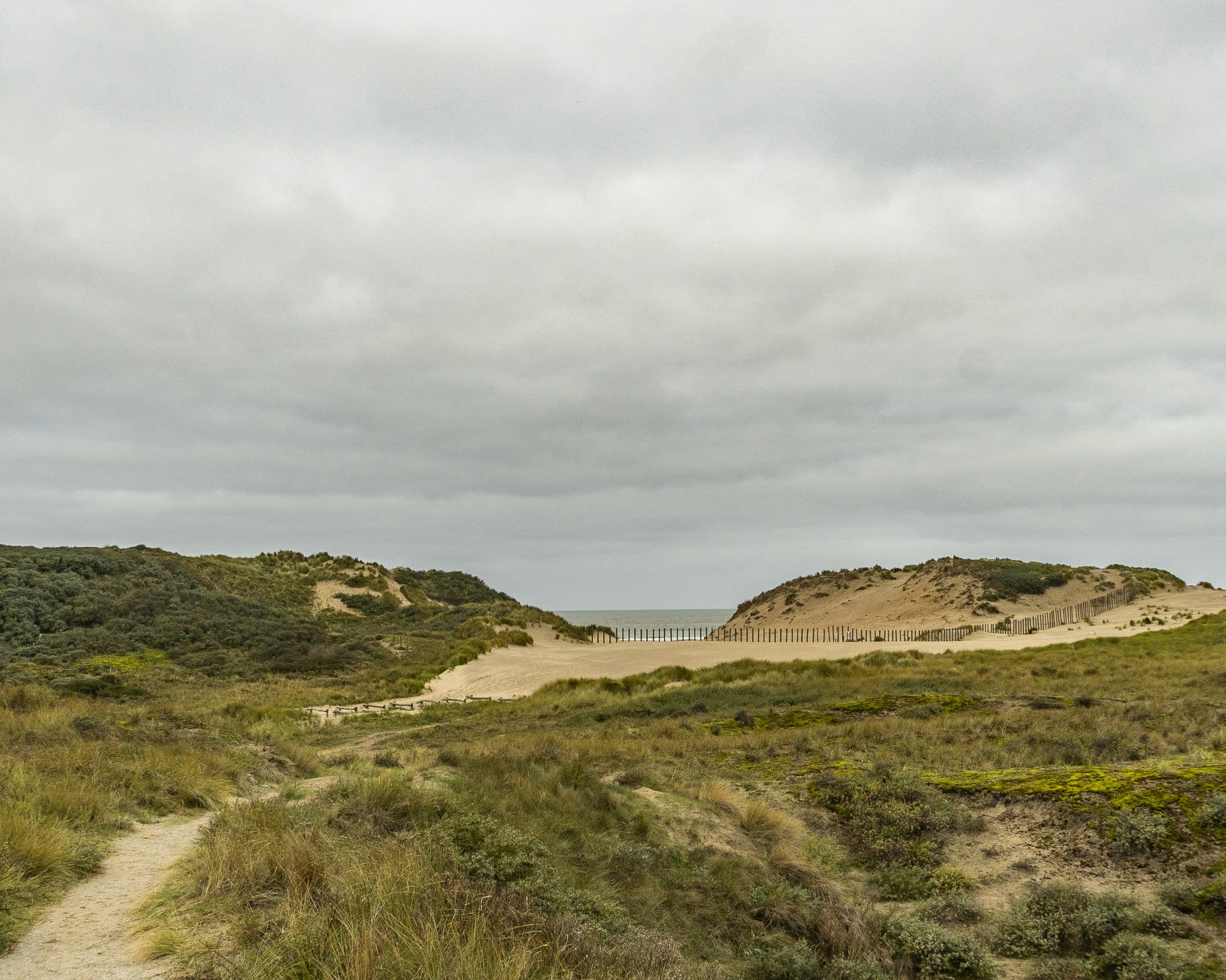 Sandy dunes and grassy fields stretch towards the sea under a cloudy sky.