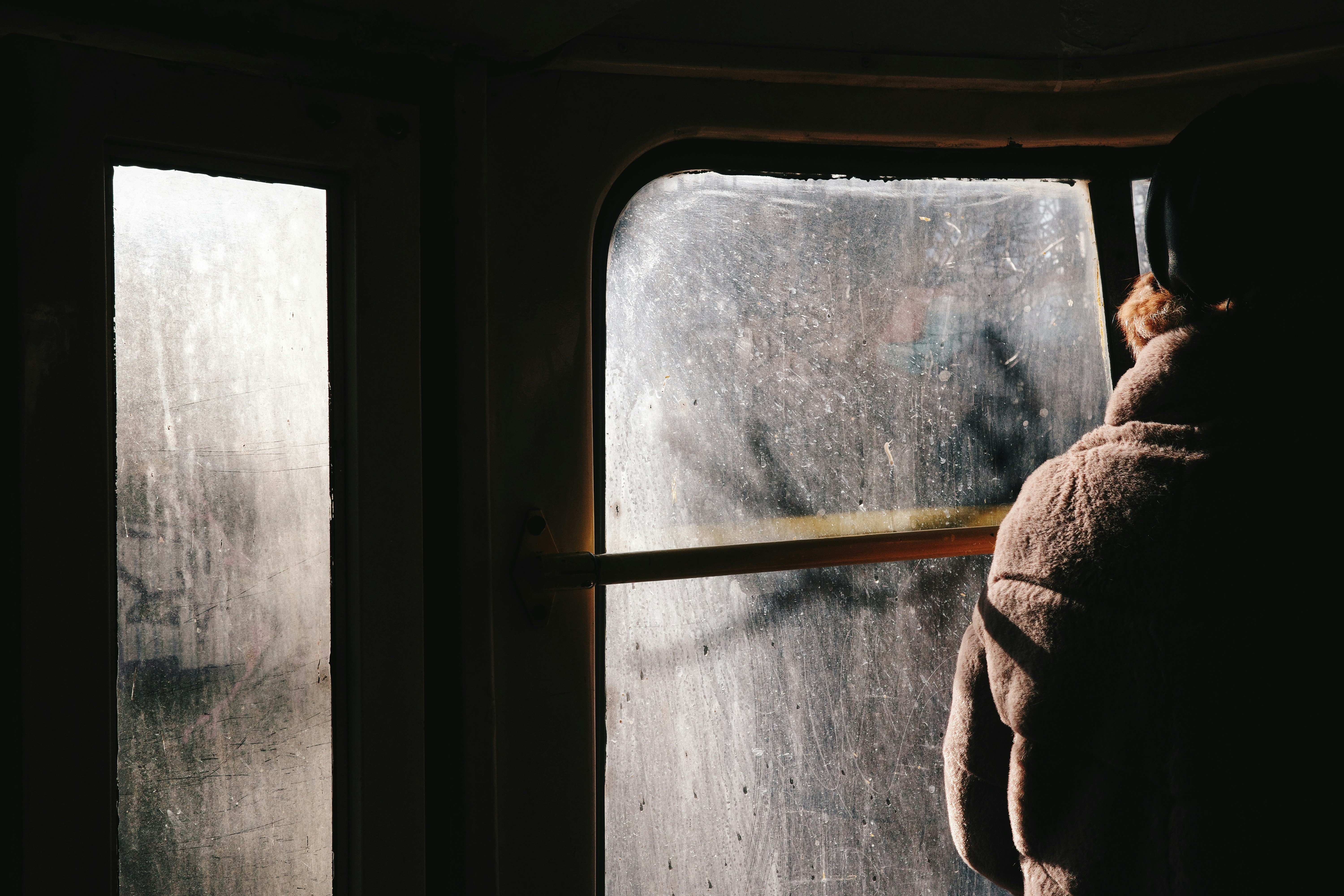 Person in a heavy coat gazes out a scratched window in dim light.