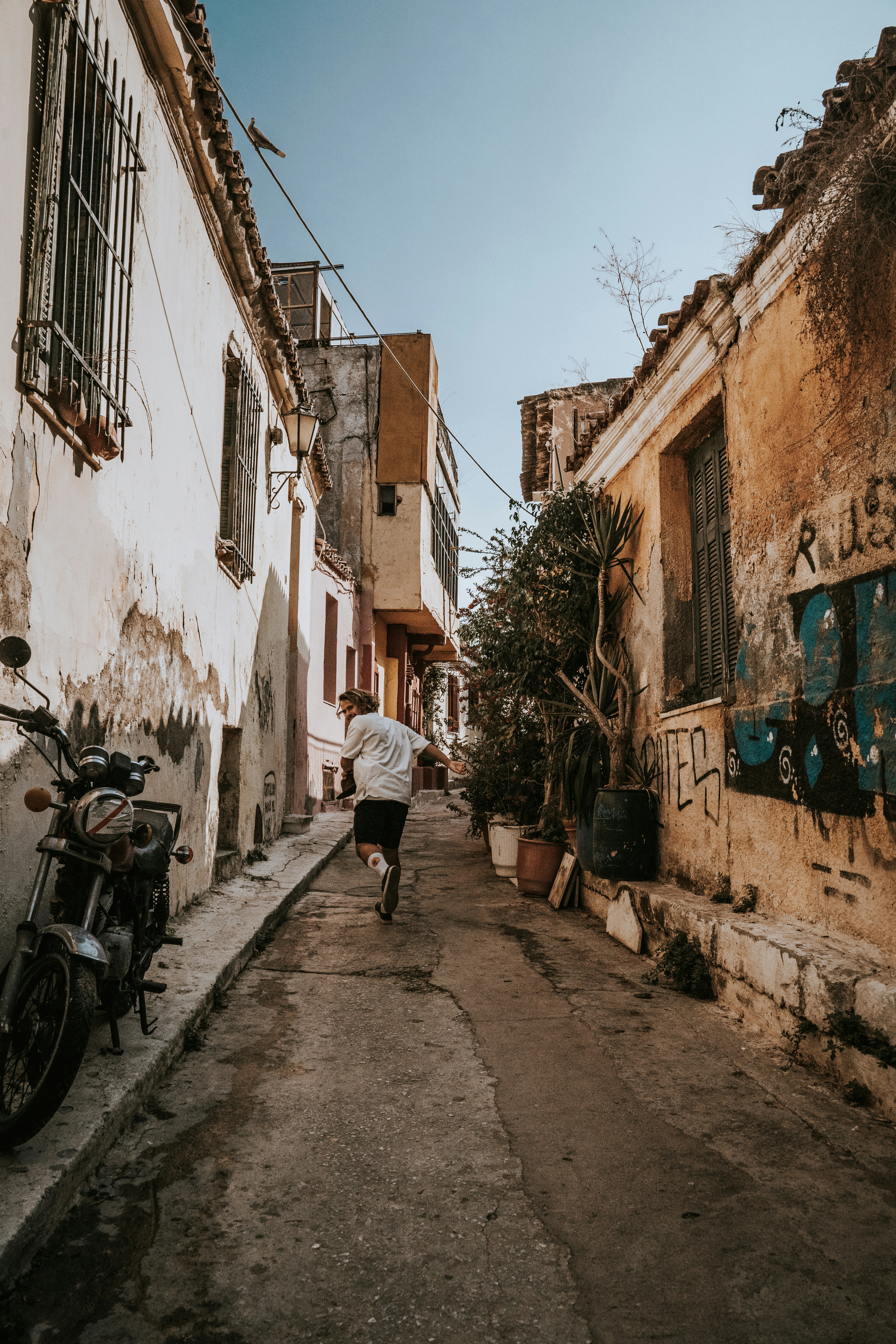 Person walking through a narrow, rustic alley with aged buildings and a clear sky.