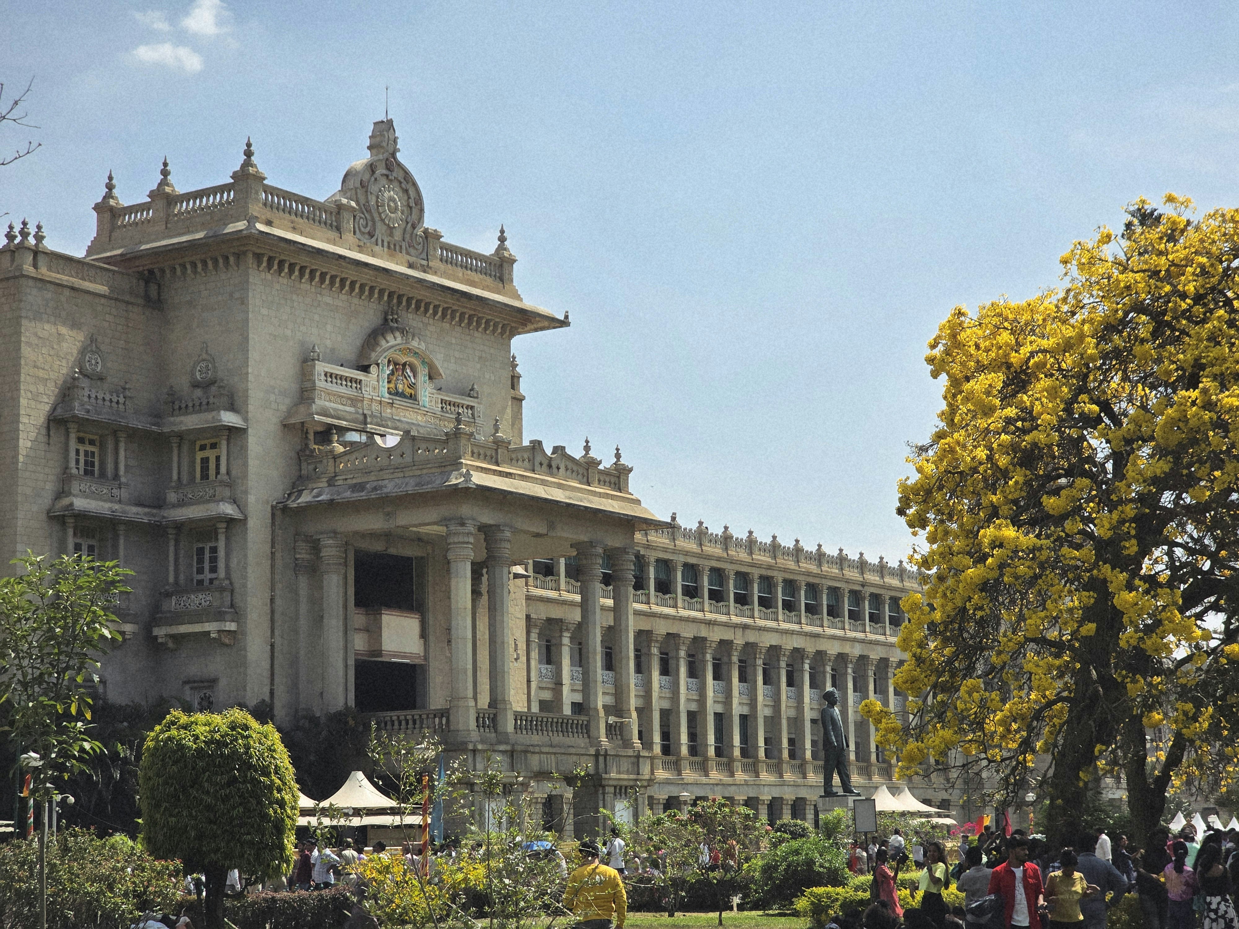 Grand neoclassical building with tall columns dominates the scene as people stroll in a crowded courtyard beneath a clear blue sky. A statue stands on a pedestal near the entrance, adding a focal point to the composition.