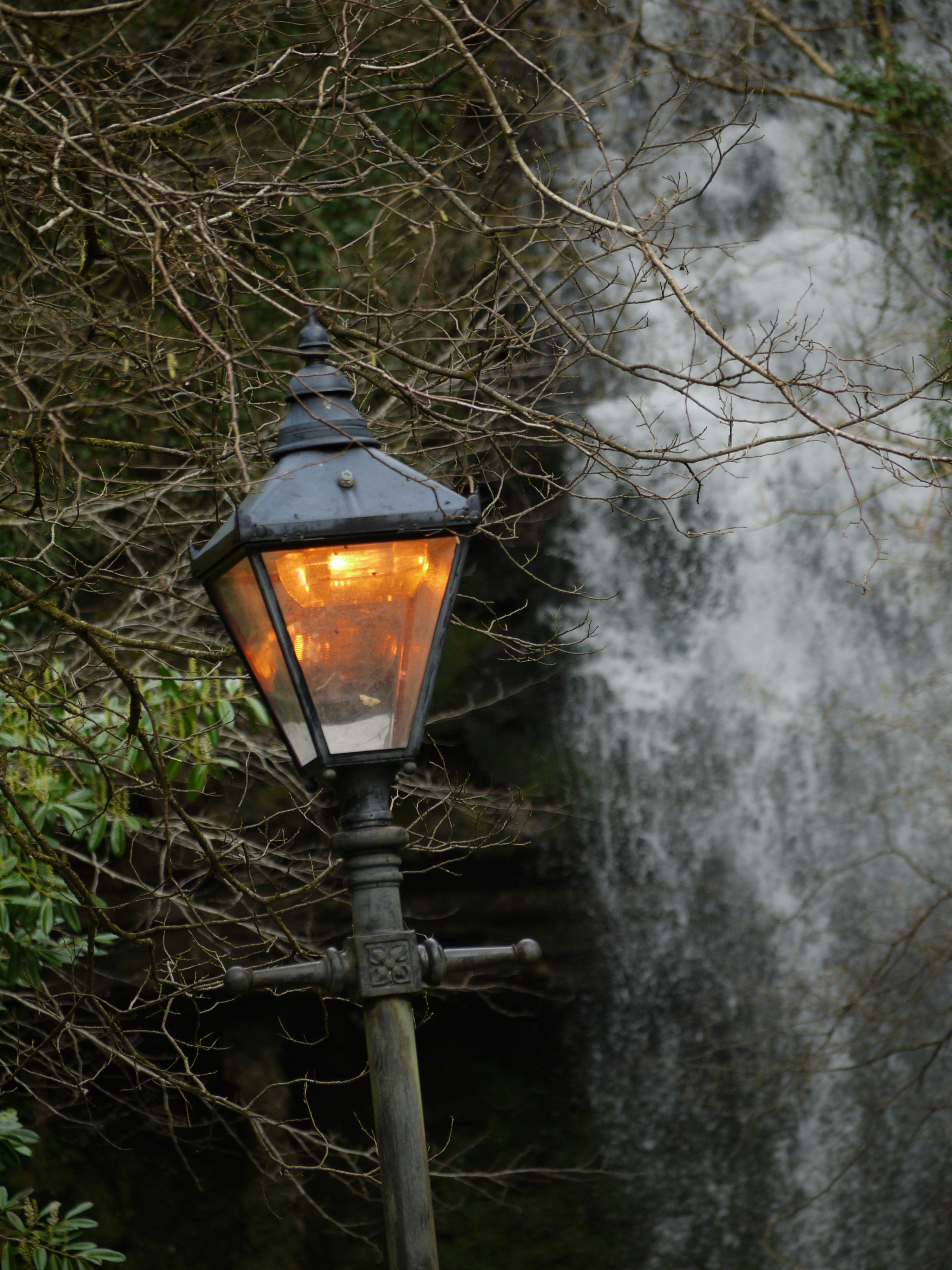 An old iron street lamp glows warmly beside bare branches, with a misty waterfall in the background.