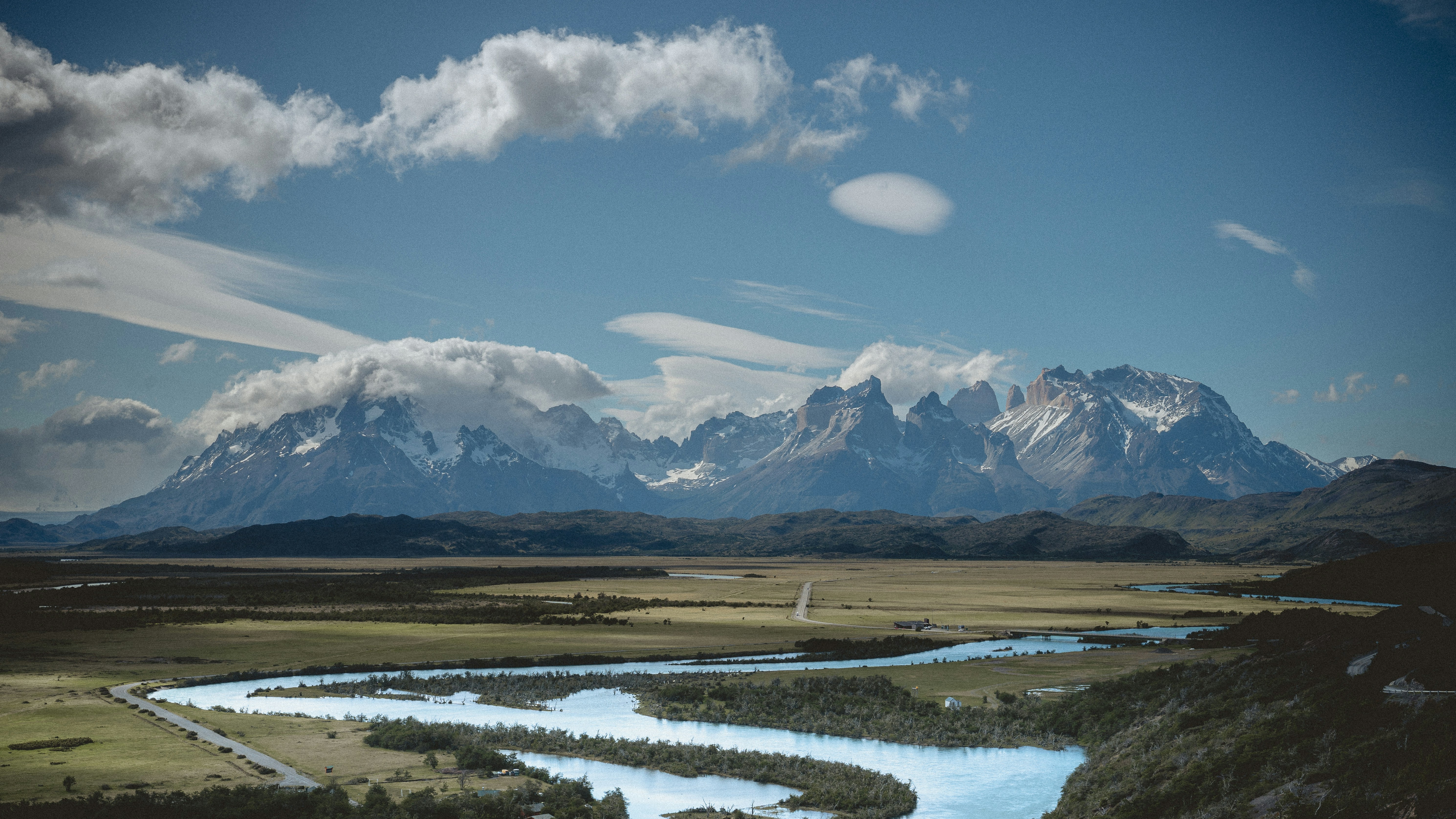 Snow-capped mountains tower over a winding river and expansive plain under a blue sky with scattered clouds.