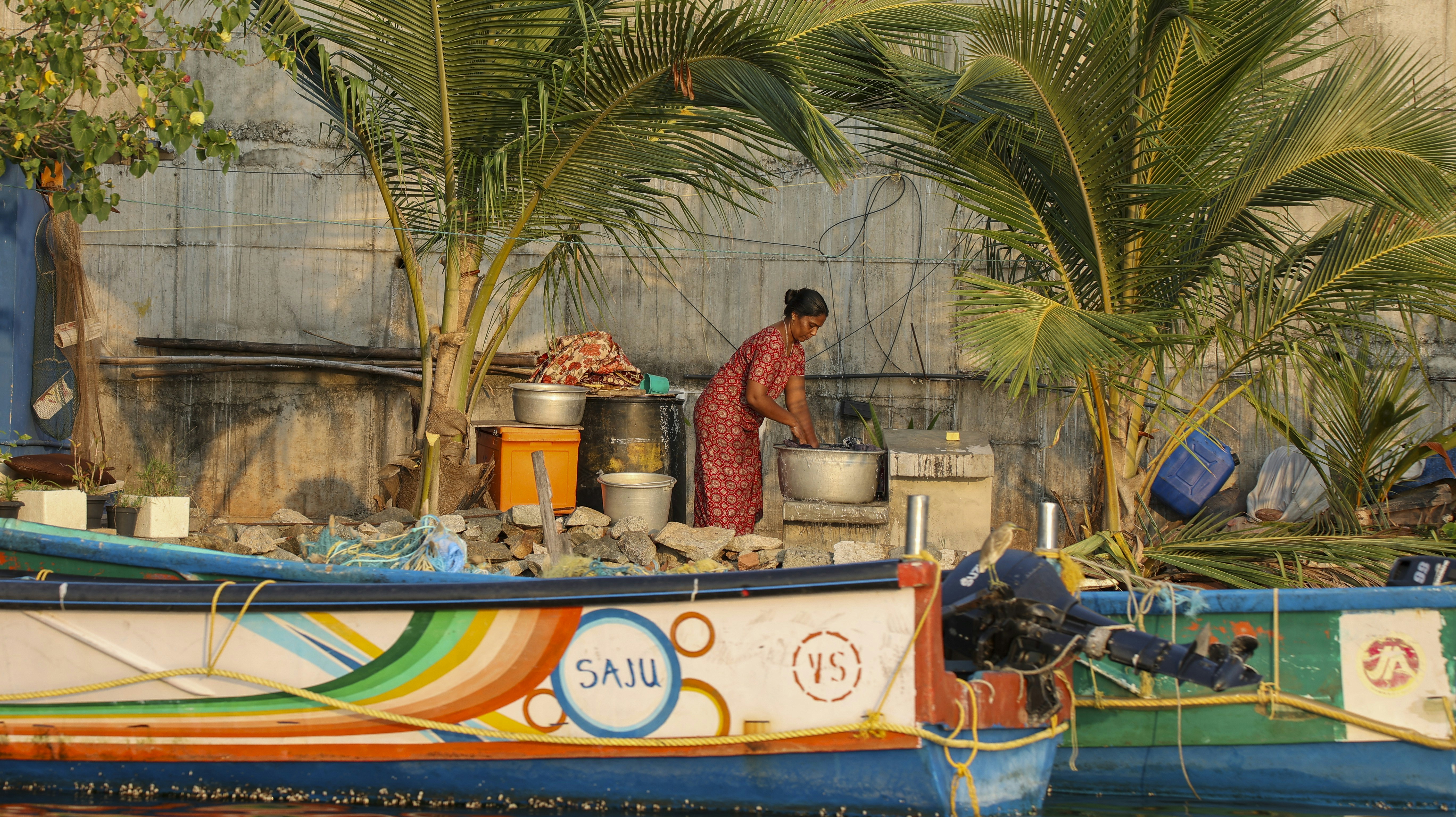 A woman washing clothes in a large basin in a fishing village, surrounded by boats, palm trees, and everyday objects. The warm golden light enhances the authenticity of this daily life scene.